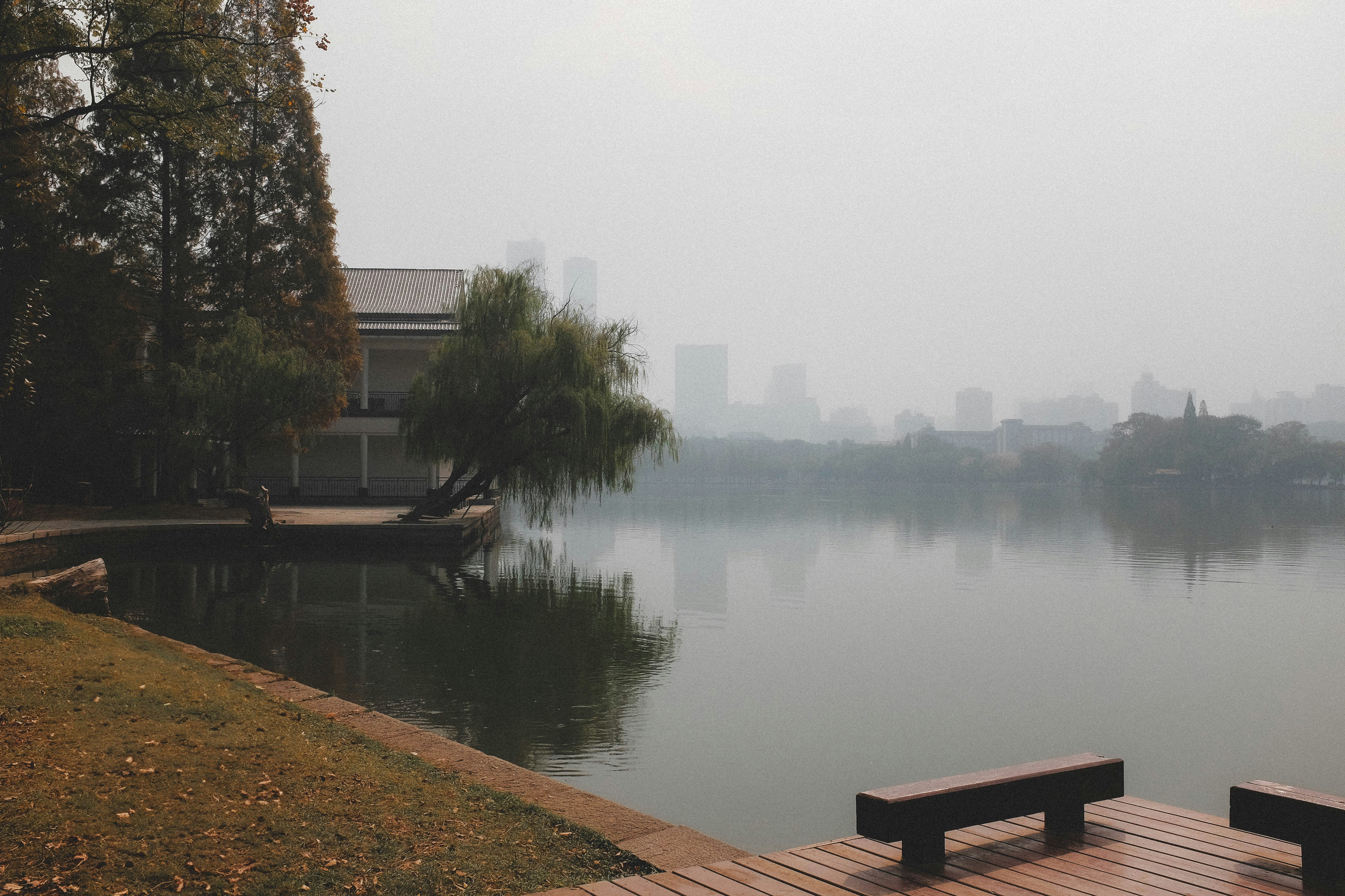 Calm lakeside scene with a wooden boardwalk, benches, and a misty city skyline in the distance.