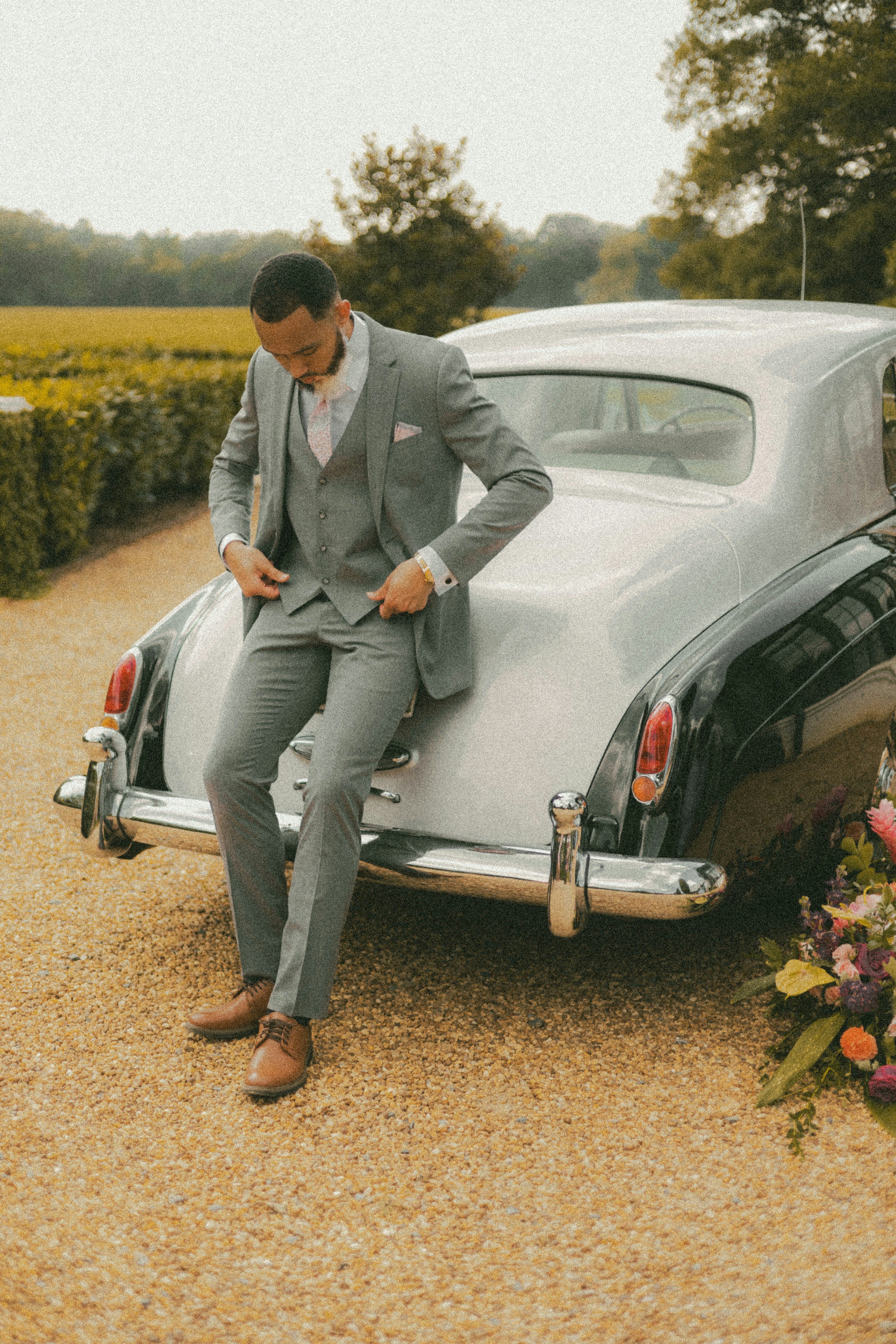 Man in a light gray suit adjusts his jacket beside a polished vintage car on a gravel path. The scene emphasizes a refined moment of preparation in an outdoor setting.