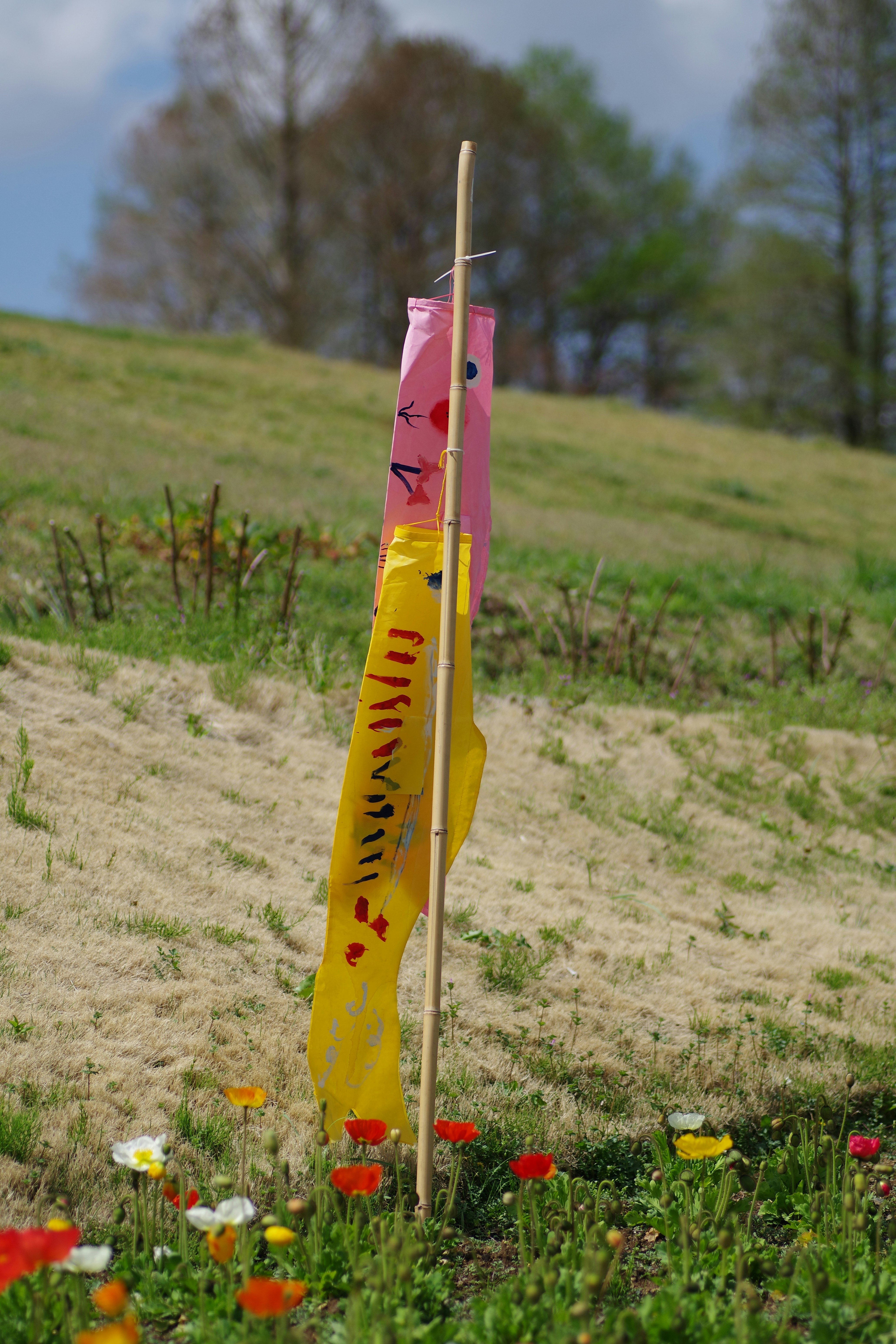 Colorful handmade flags fluttering on a stick amidst a vibrant flower bed in a sunlit landscape.