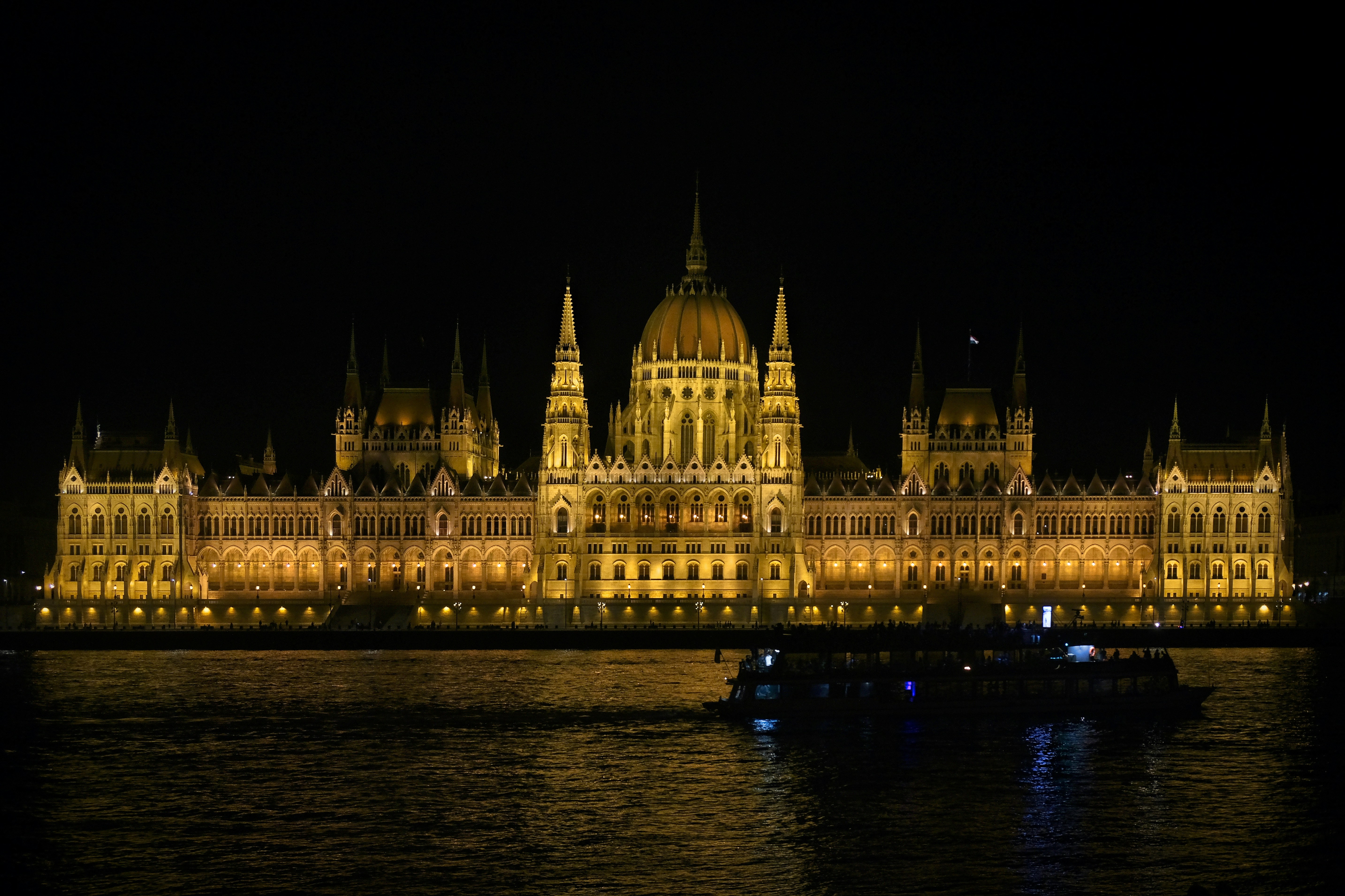 A breathtaking nighttime capture of the Hungarian Parliament Building in Budapest, illuminated with golden lights and reflected in the tranquil waters of the Danube River. The Gothic Revival architecture glows majestically against the pitch-black sky, while a silhouetted boat adds a touch of life and depth to the foreground.