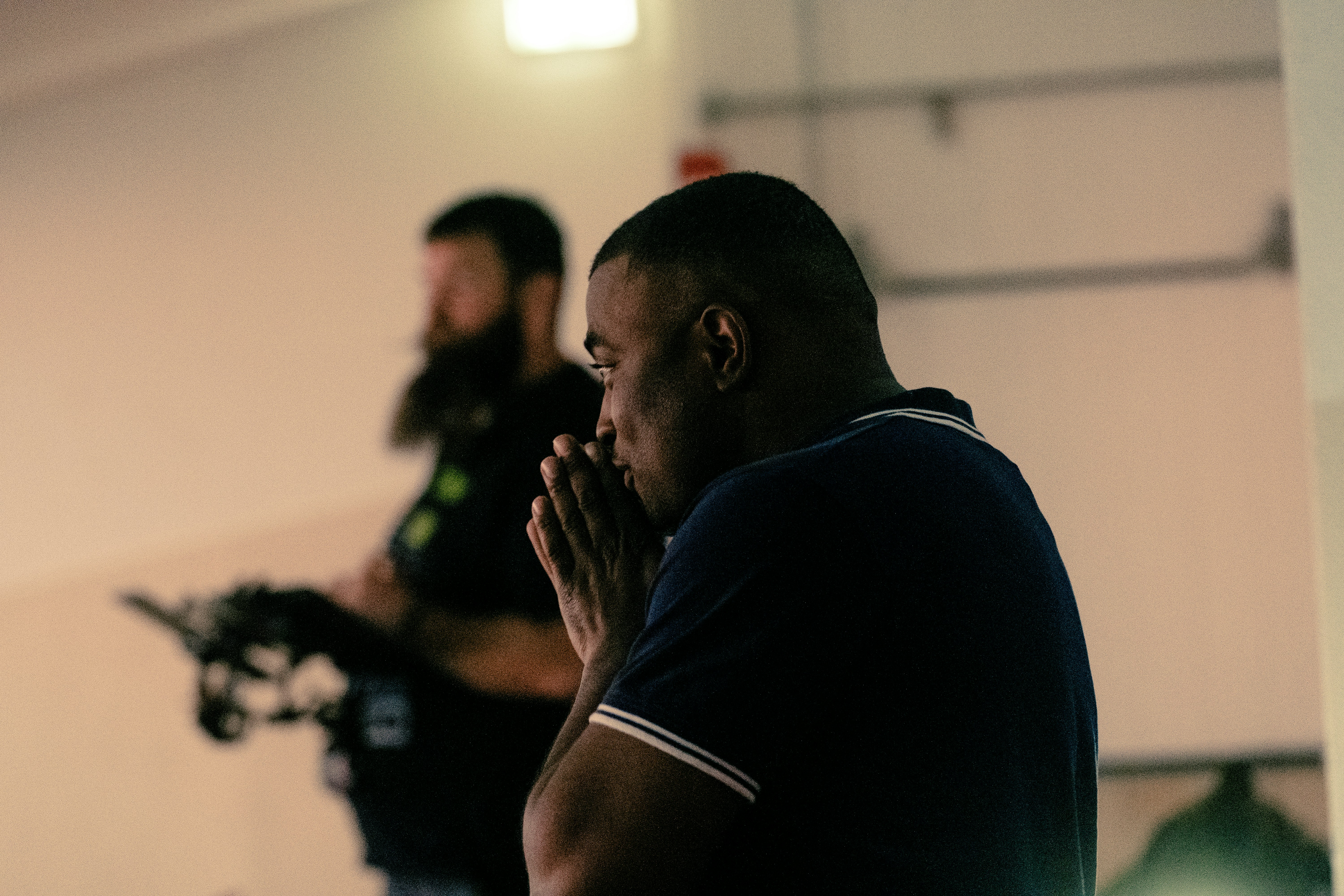 A focused man with clasped hands stands in contemplation, while a bearded figure operates a camera in the background. The scene captures a moment of tension and preparation.