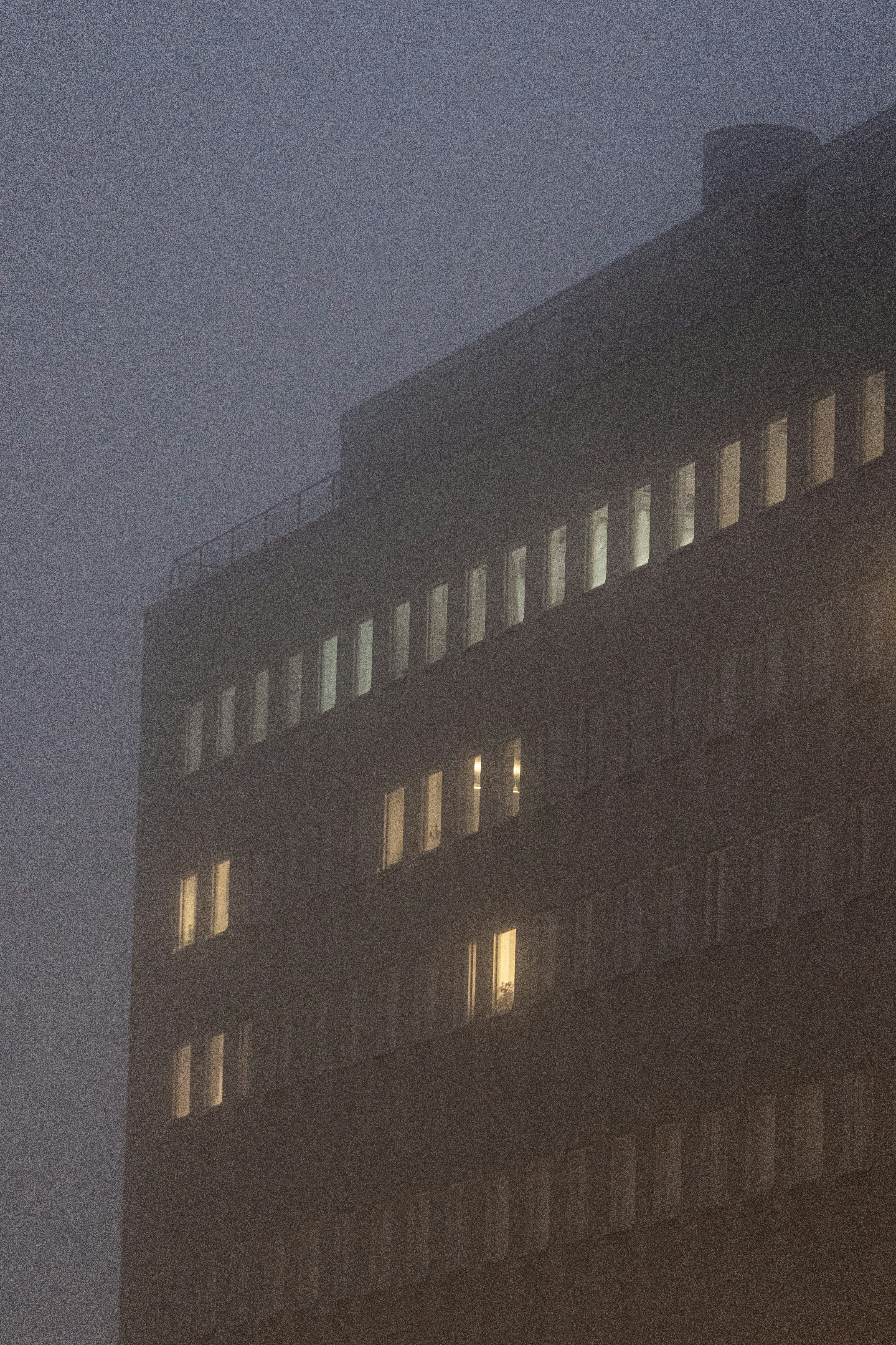 Illuminated windows of a building emerge from a dense fog, creating an atmosphere of mystery and intrigue.