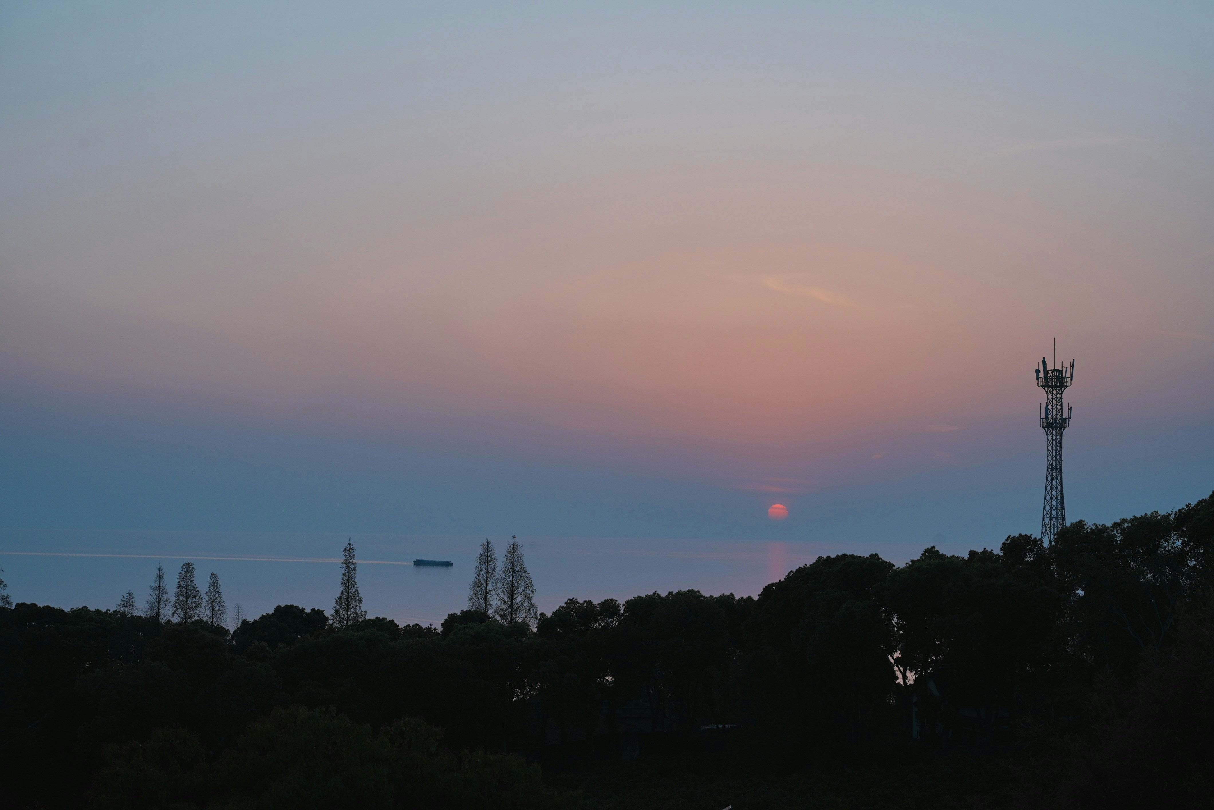 Pastel sunset over a treeline with a distant cell tower on the right, silhouetted against a calm horizon.