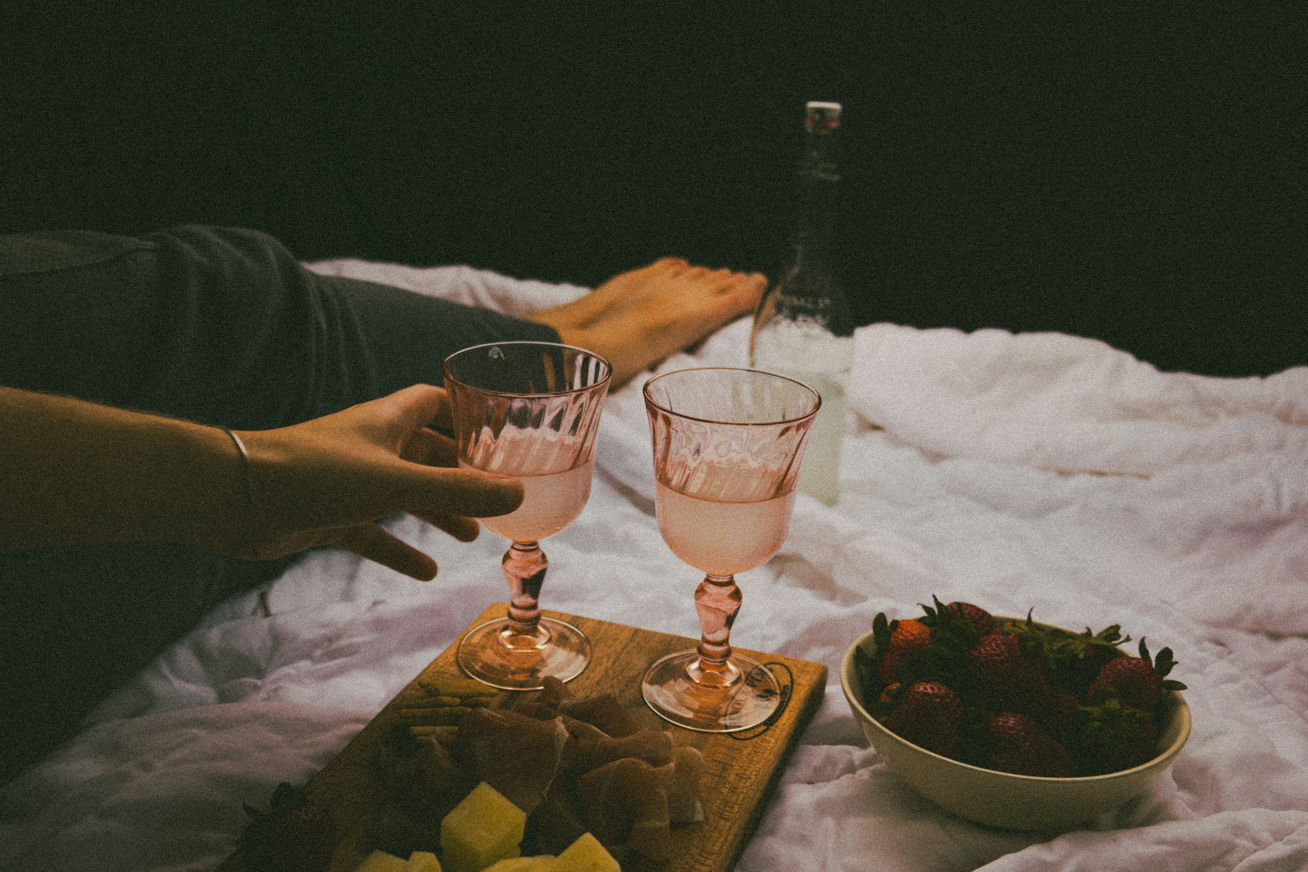 Two elegant glasses of cocktail are raised in a cozy setting, accompanied by a platter of fruits and a bottle of drink. The scene evokes a sense of relaxation and indulgence.
