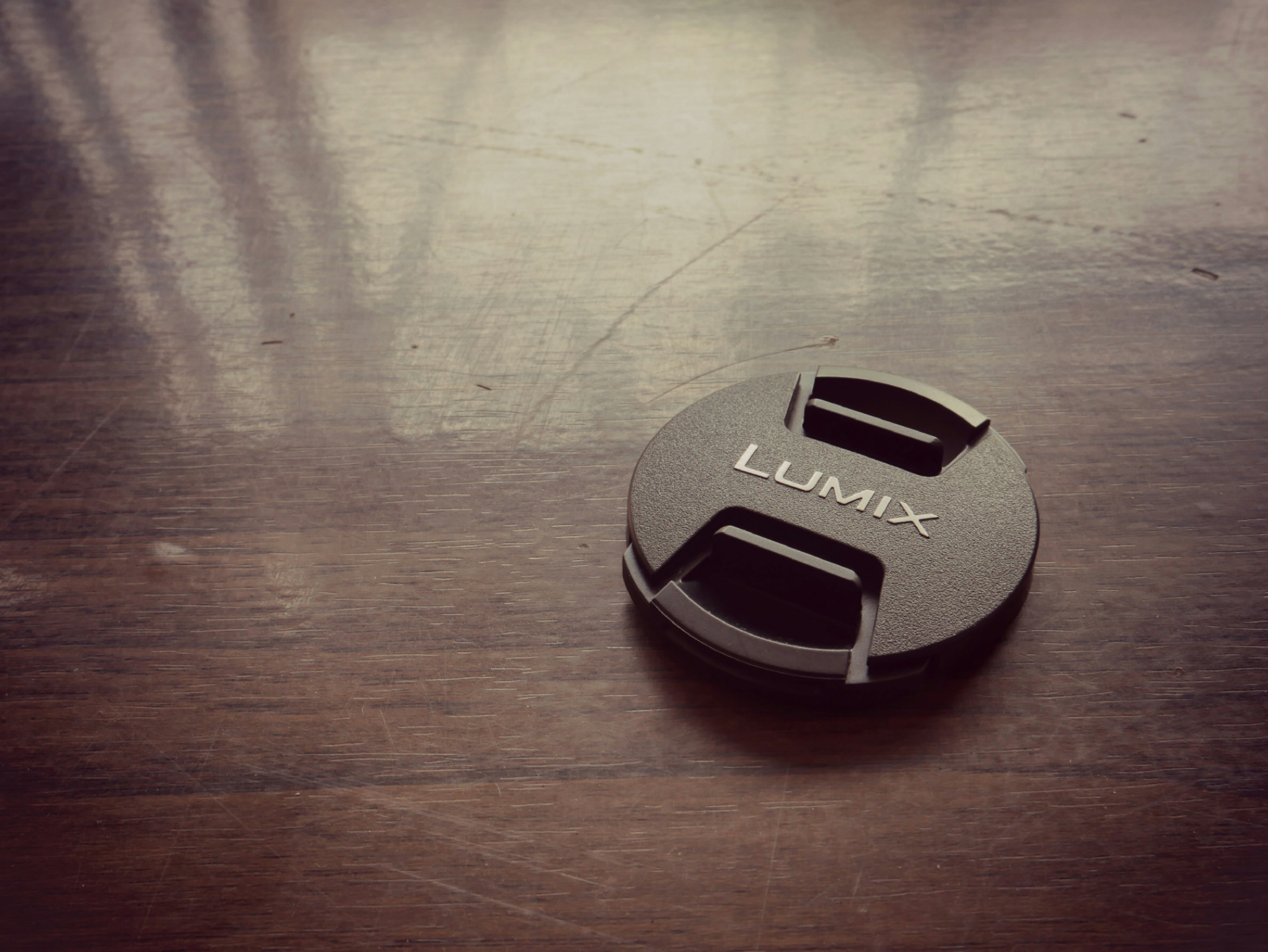 A Panasonic Lumix lens cap resting on a wooden surface, showcasing its sleek design and branding. The soft lighting accentuates the textures of the cap and the table beneath.