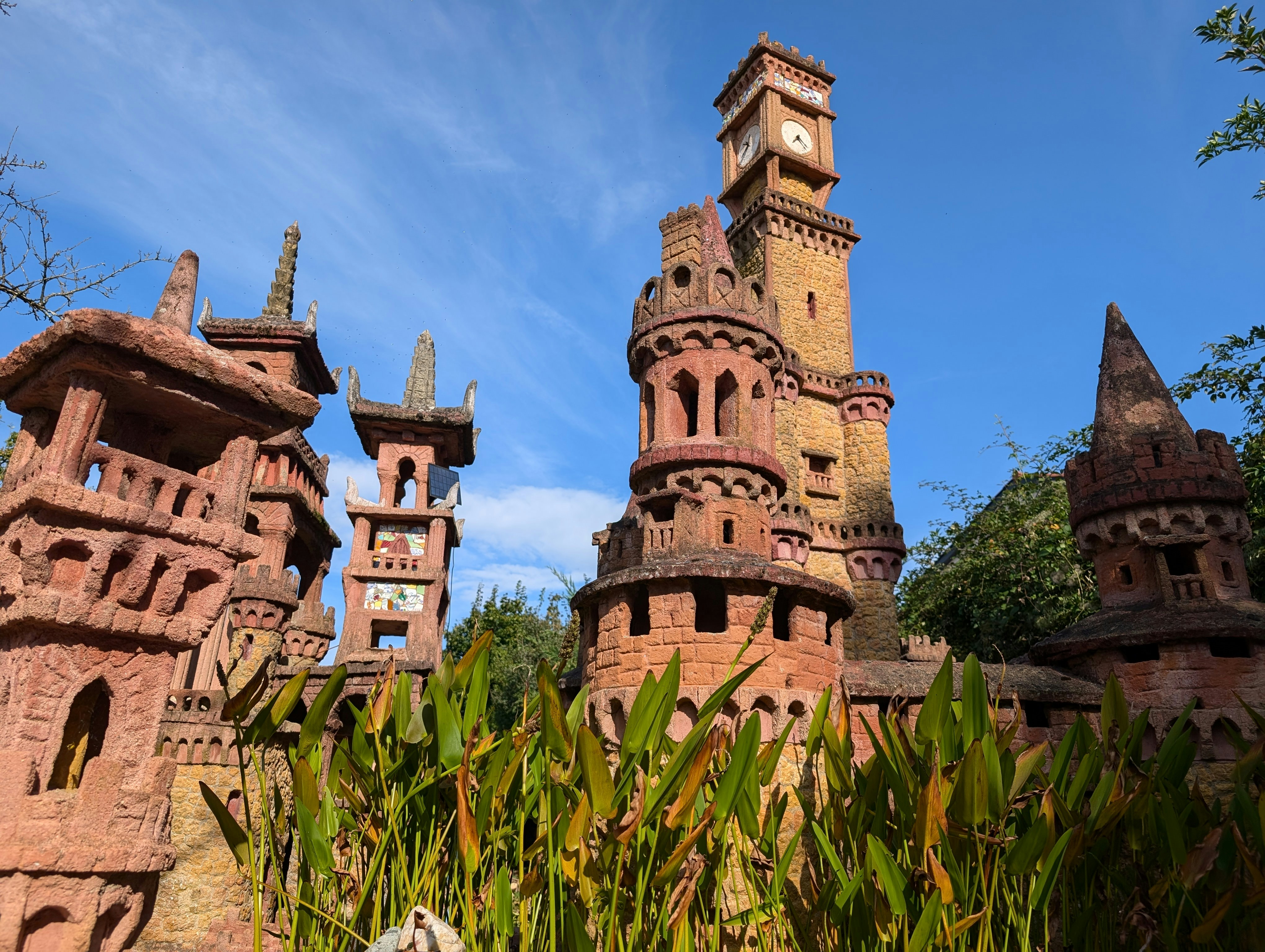 Towering brick structures rise behind lush tropical ferns, with a bright blue sky above; a decorative, fortress-like landscape scene featuring weathered red masonry and intricate architectural details.