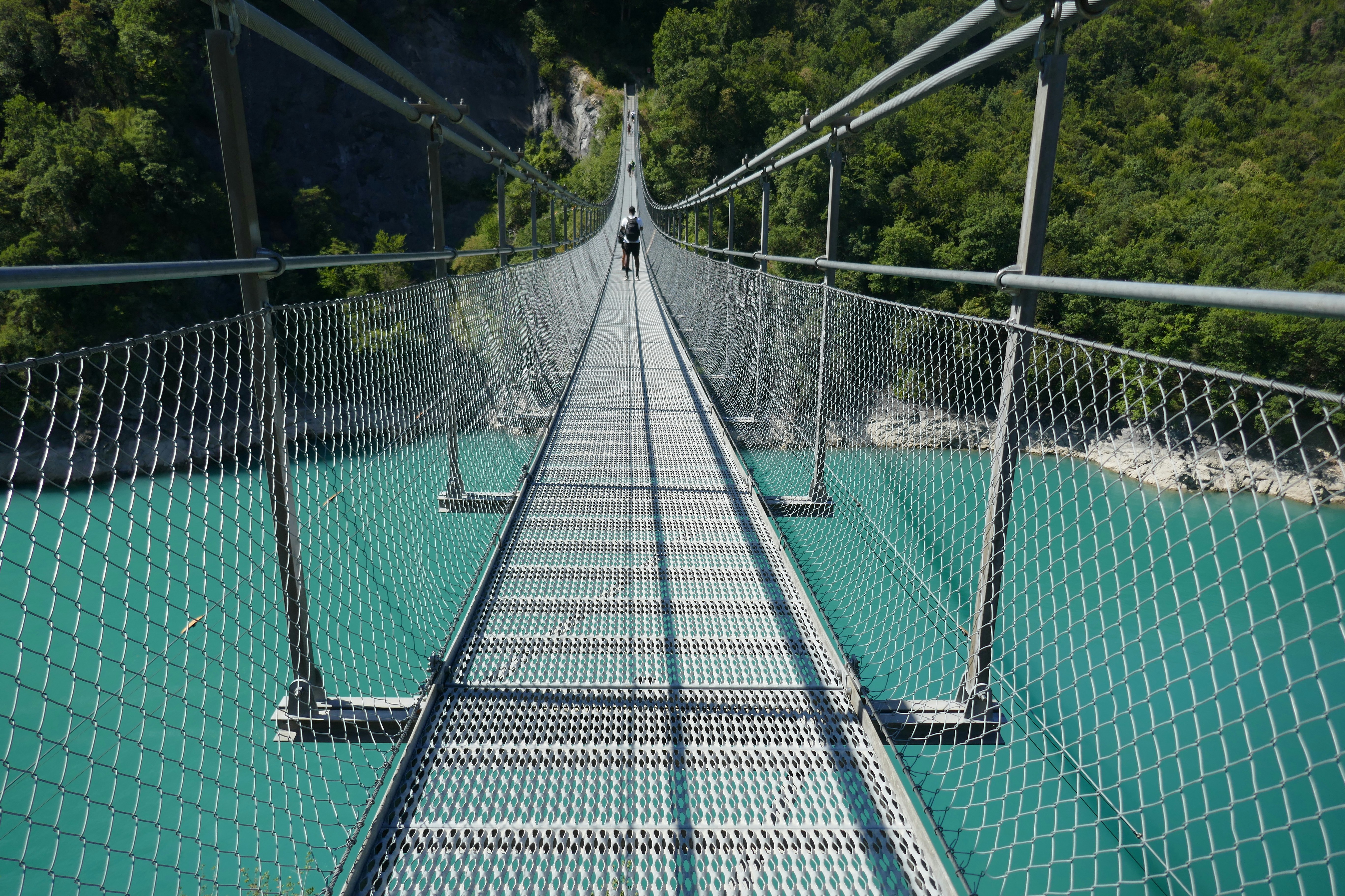 Photograph of a suspension bridge spanning a turquoise river, drawing the eye toward distant hikers at the far end. The metal walkway and safety mesh contrast with vivid water and surrounding forest.