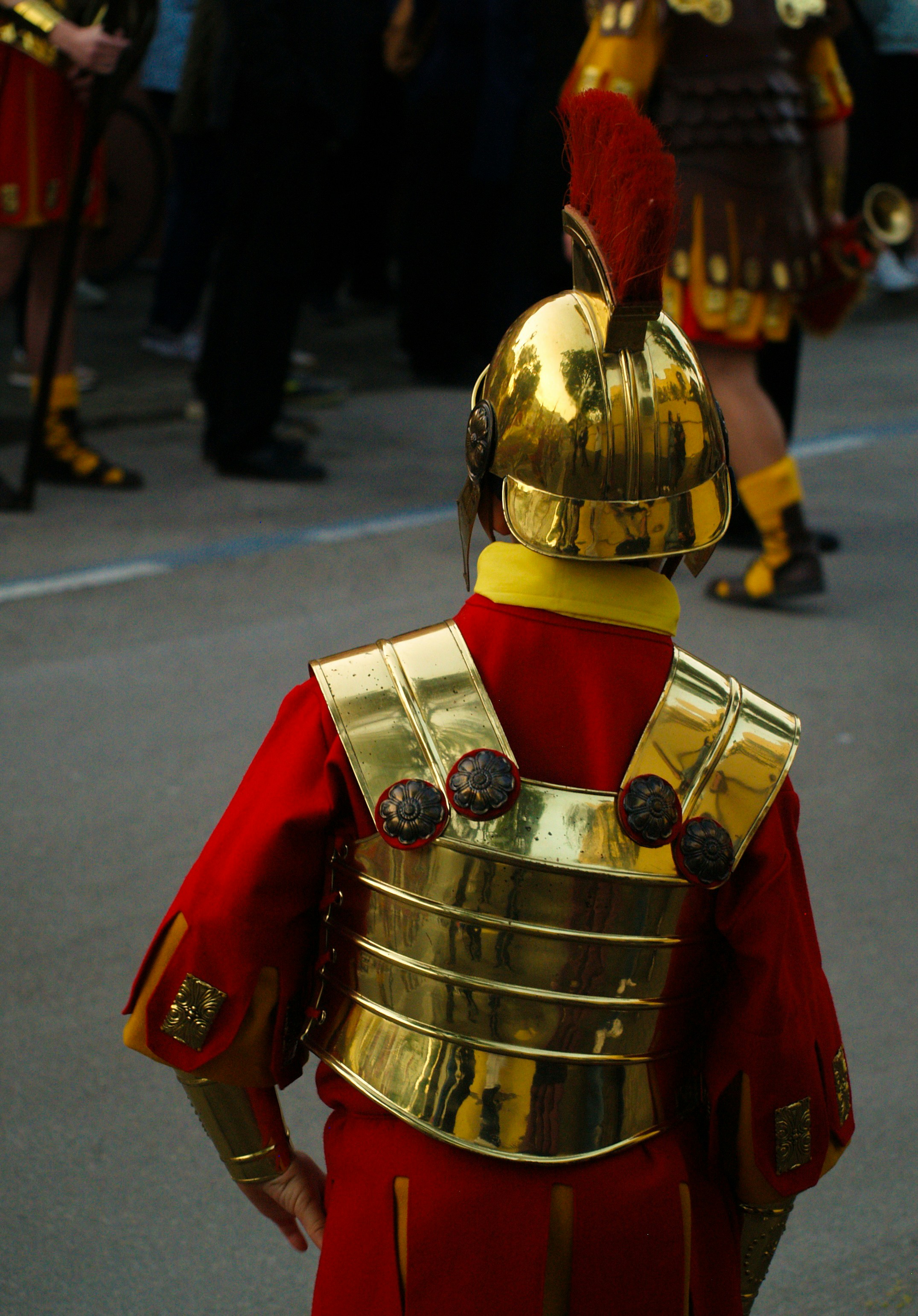 A Roman soldier in ornate golden armor stands with his back to the viewer, showcasing intricate details against a blurred crowd. The vibrant red uniform contrasts with the muted background.