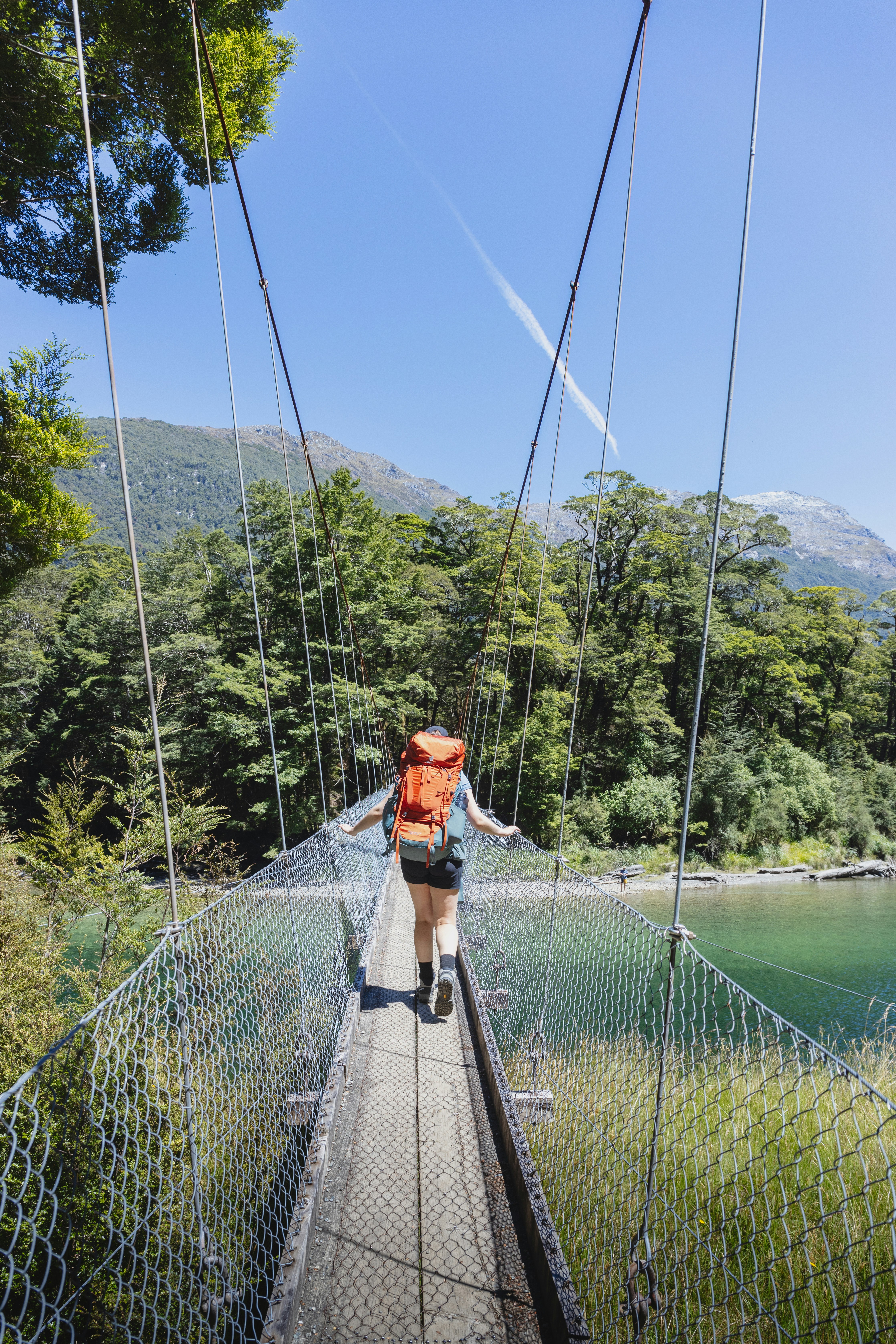 Hiker with an orange backpack traverses a suspension bridge above a lush forest and clear river under a bright blue sky.