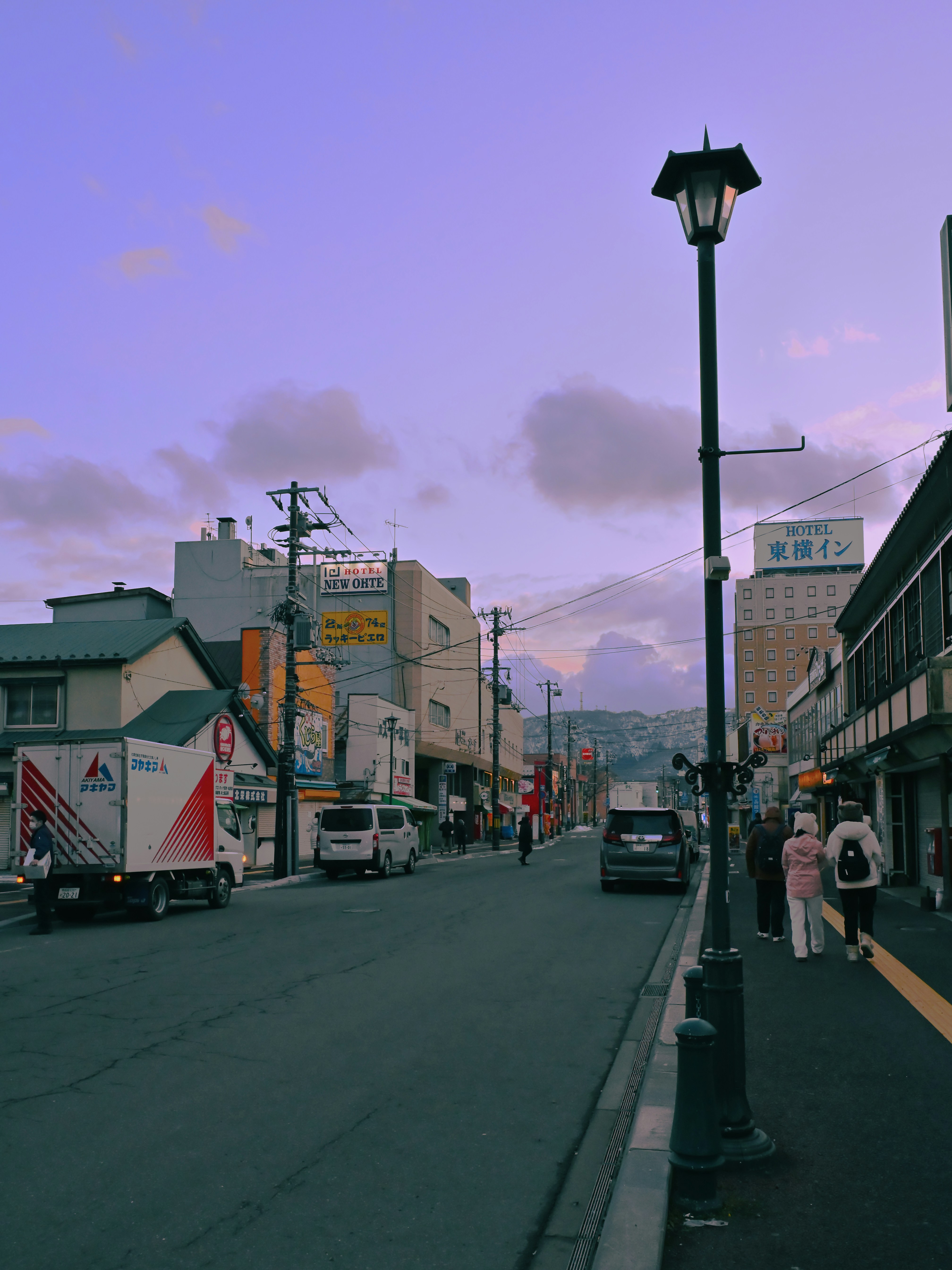 A serene street scene at dusk, featuring a mix of vehicles and pedestrians amidst charming storefronts and soft twilight hues.