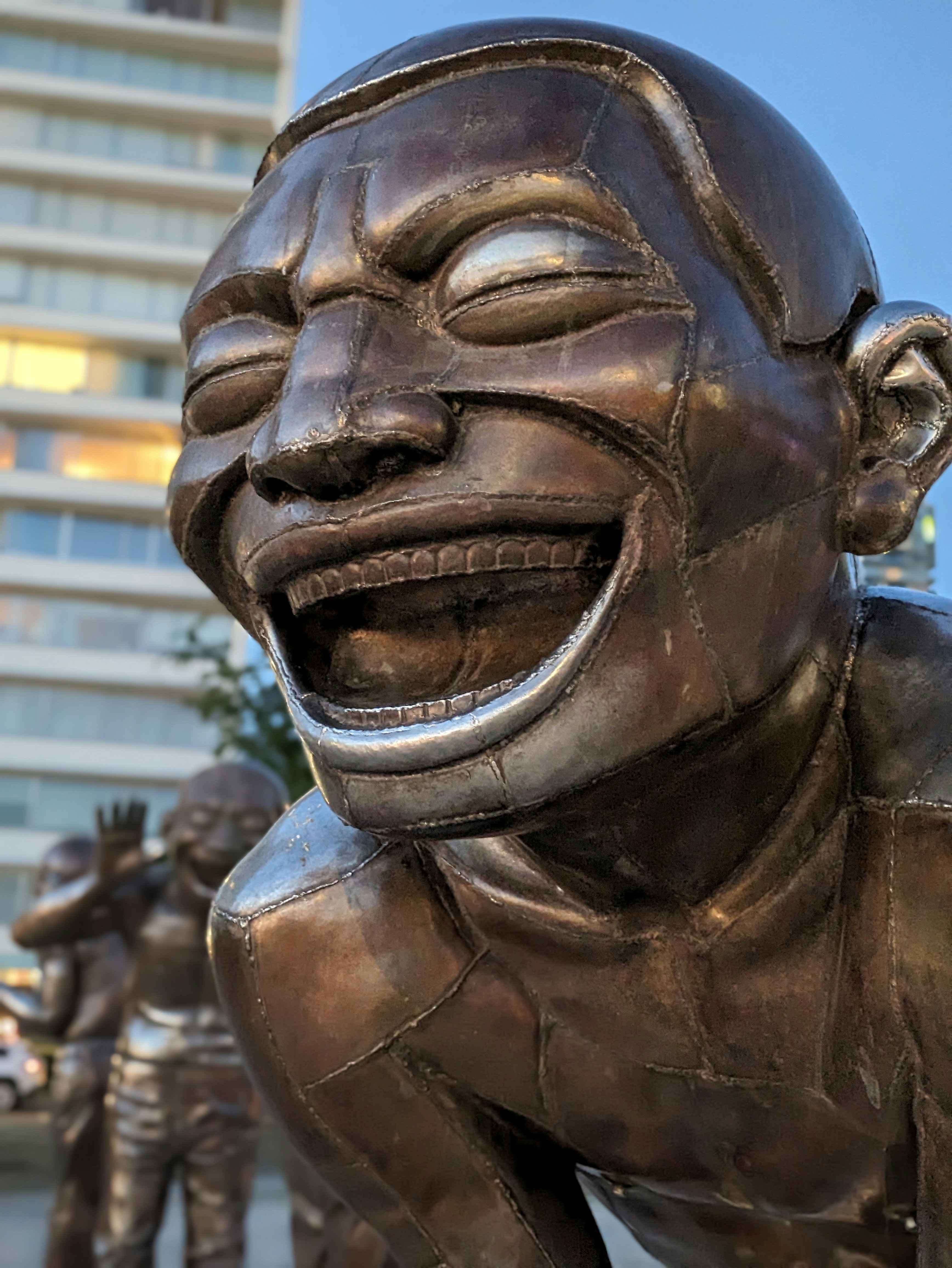 Close-up of a bronze laughing sculpture with a modern city backdrop.