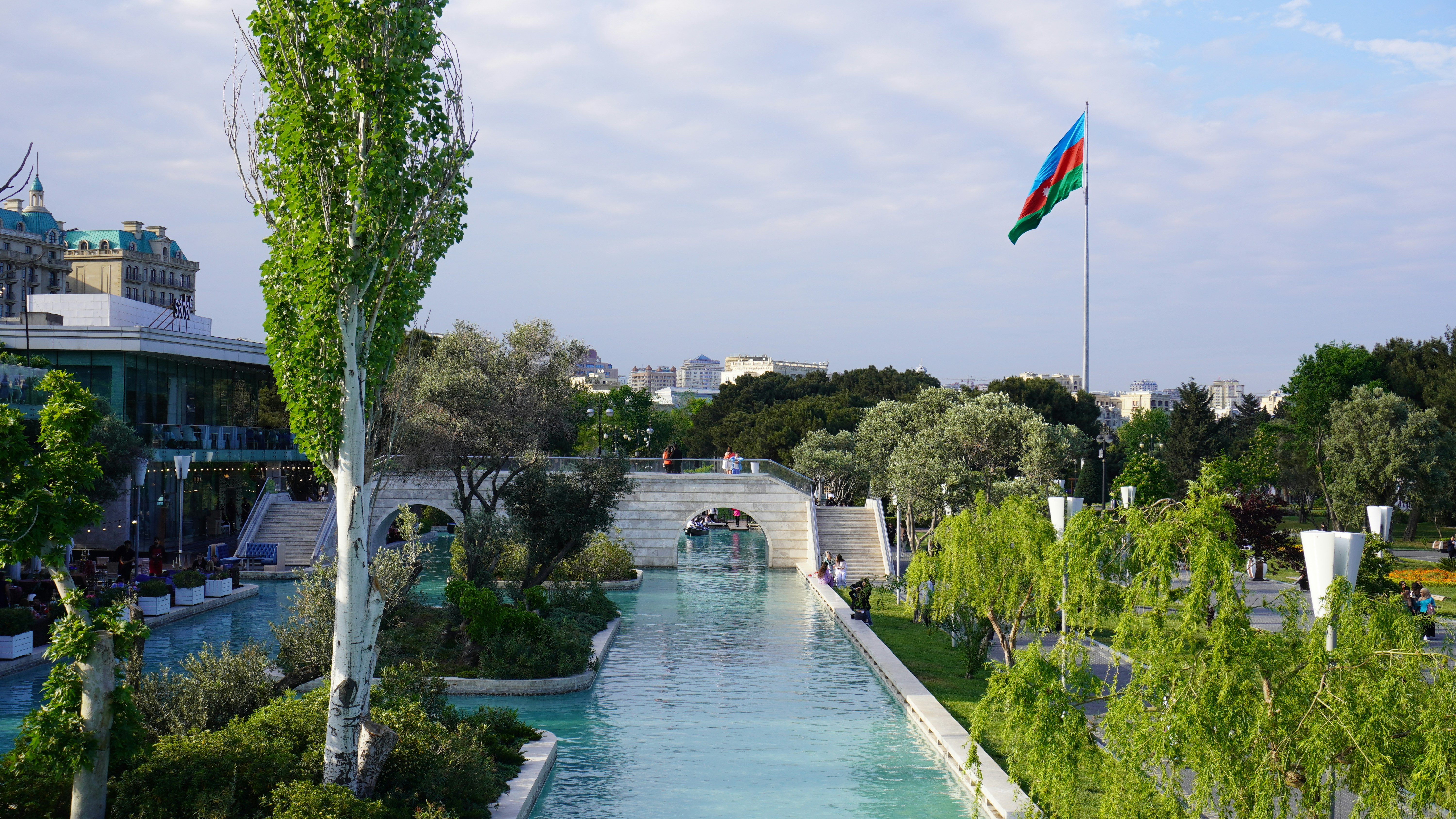 A tranquil park scene along a waterway in Baku, featuring lush greenery and a prominent flag waving in the breeze.