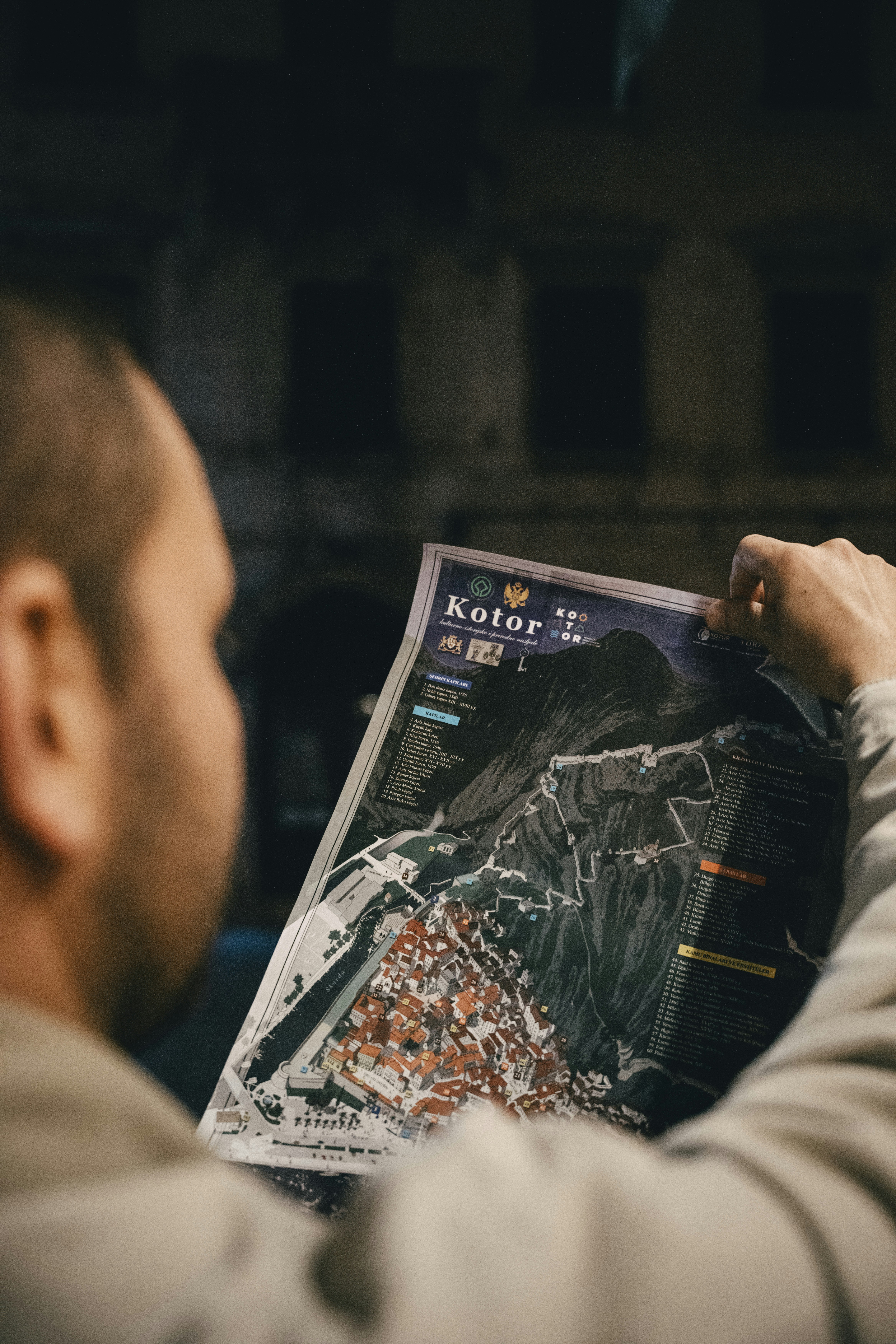A person holds a detailed map of Kotor at night, viewed from the side. The map dominates the frame as the focal point.