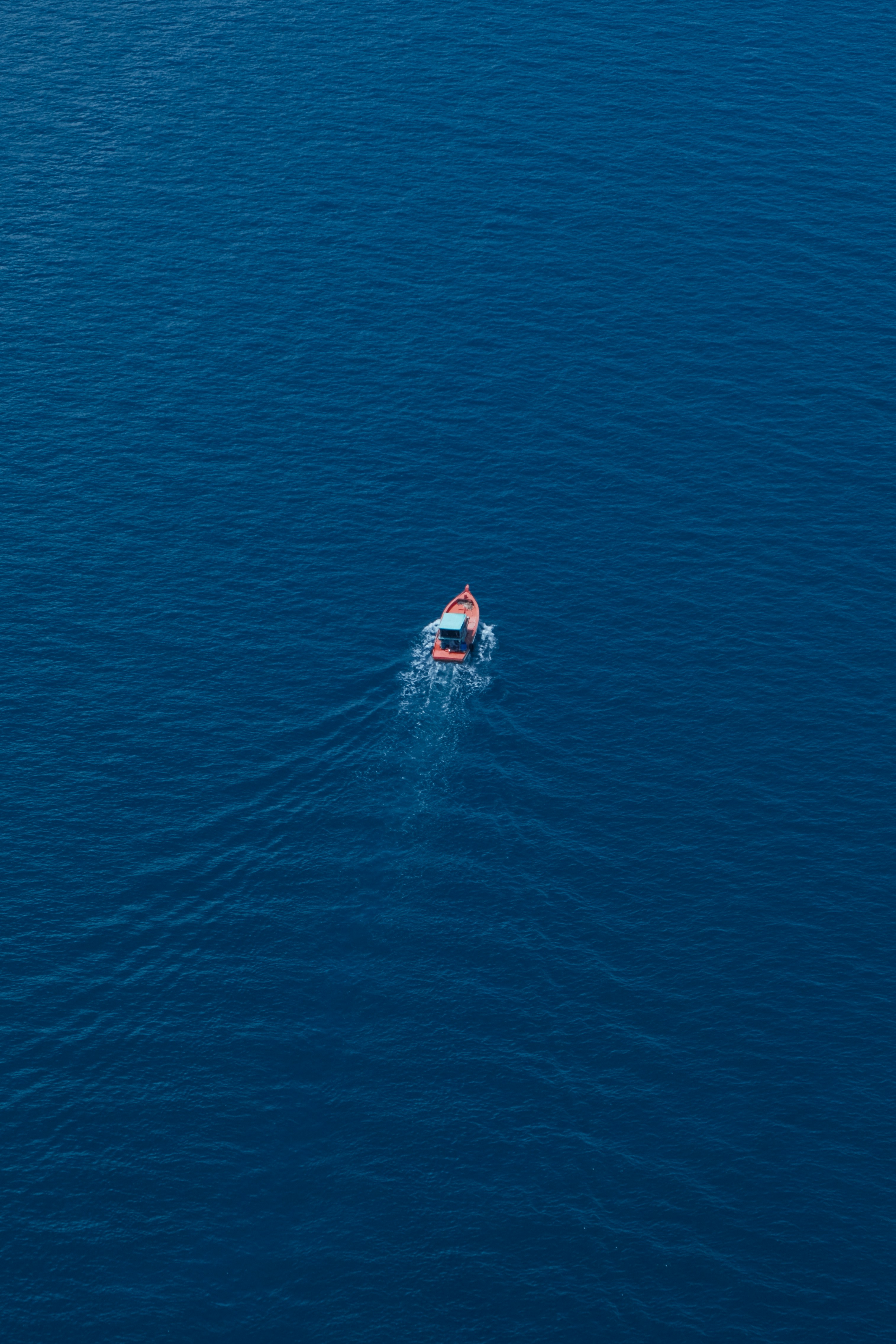 A small red boat glides through a vast expanse of deep blue water, leaving gentle ripples in its wake.