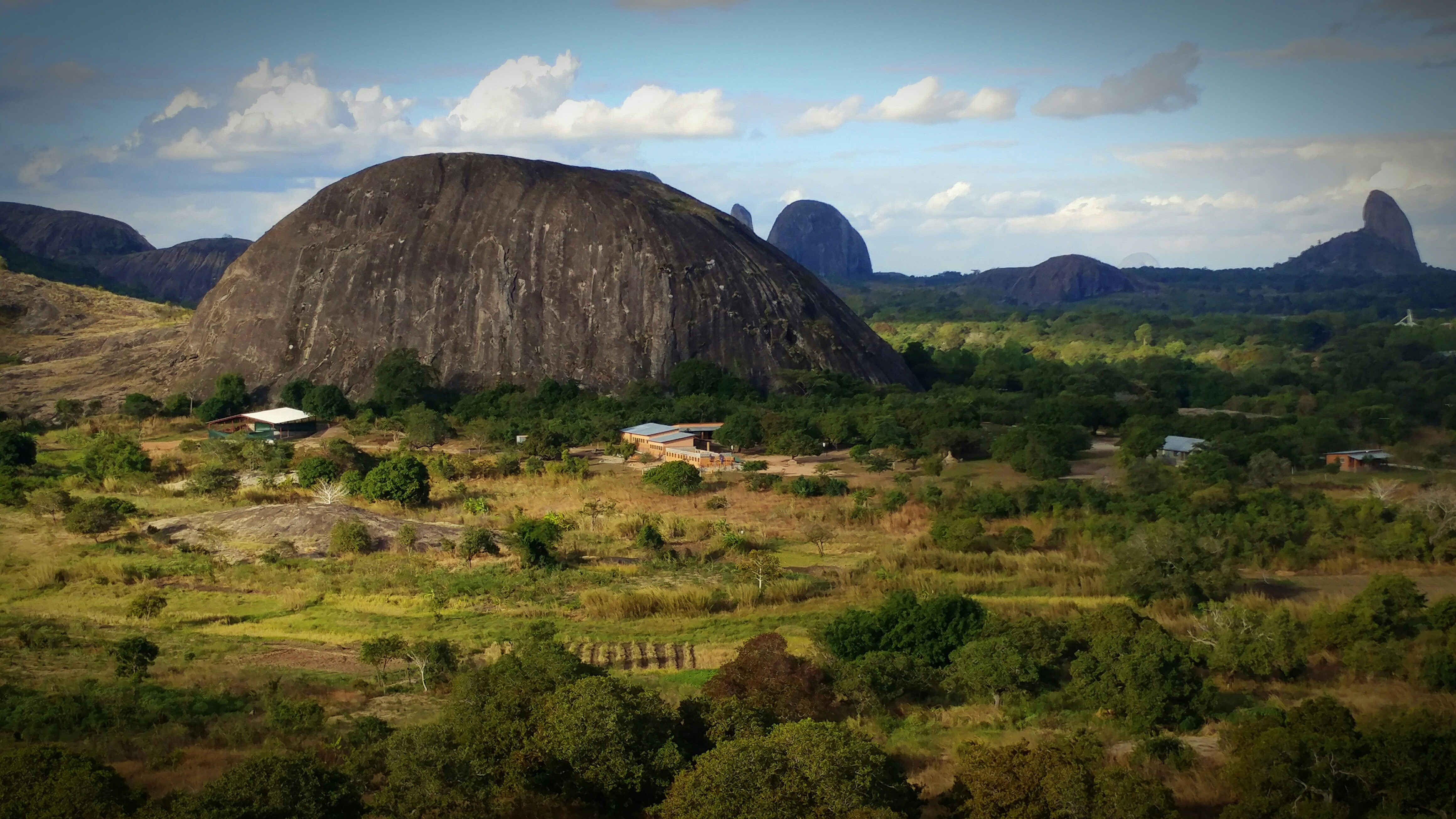 A large, rounded rock formation dominates the landscape, surrounded by lush greenery and distant hills under a clear sky.