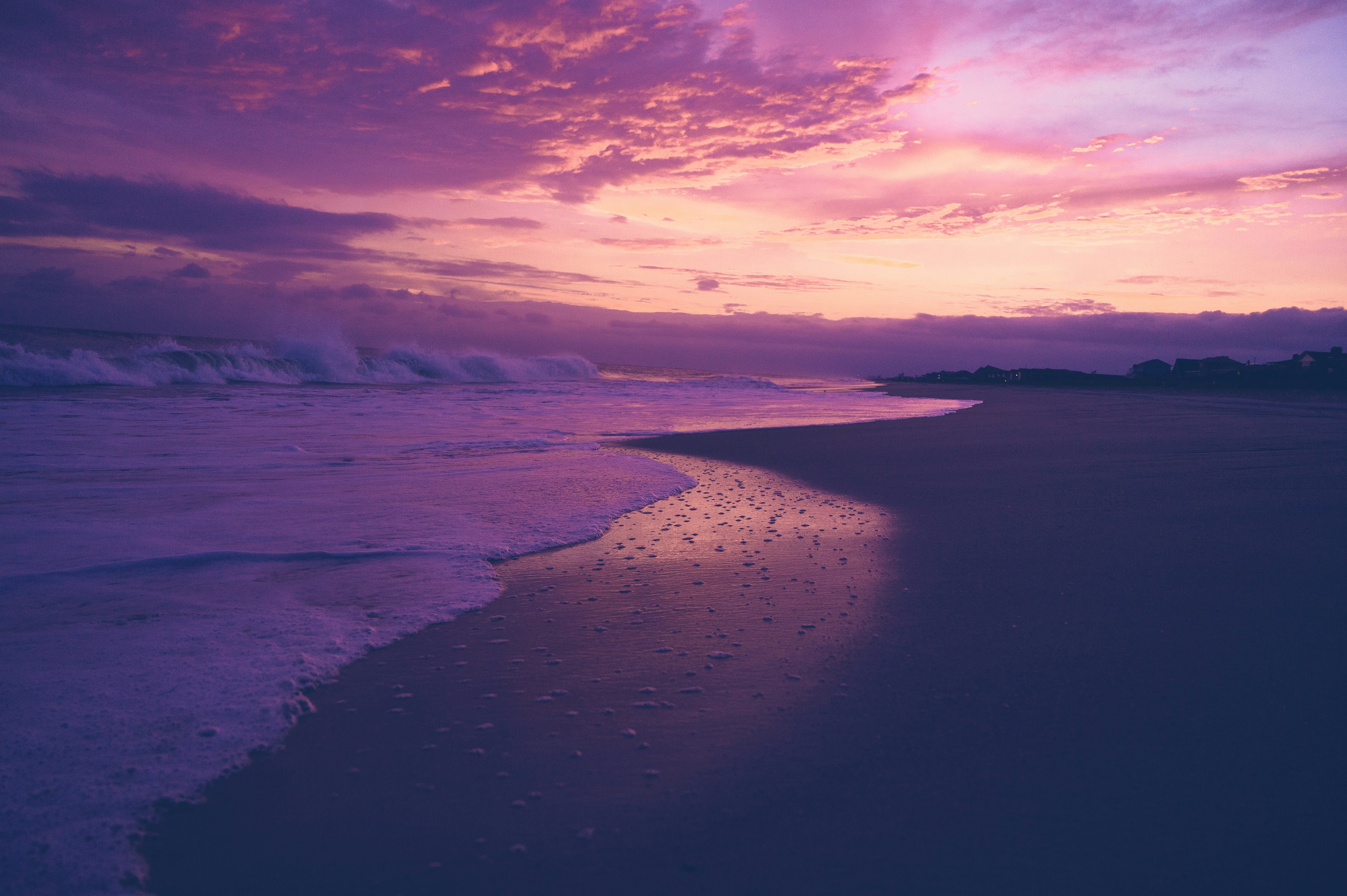 Gentle waves kiss the sandy beach under a vibrant twilight sky, where hues of purple and pink meld together. The scene captures the tranquil beauty of nature's transition from day to night.