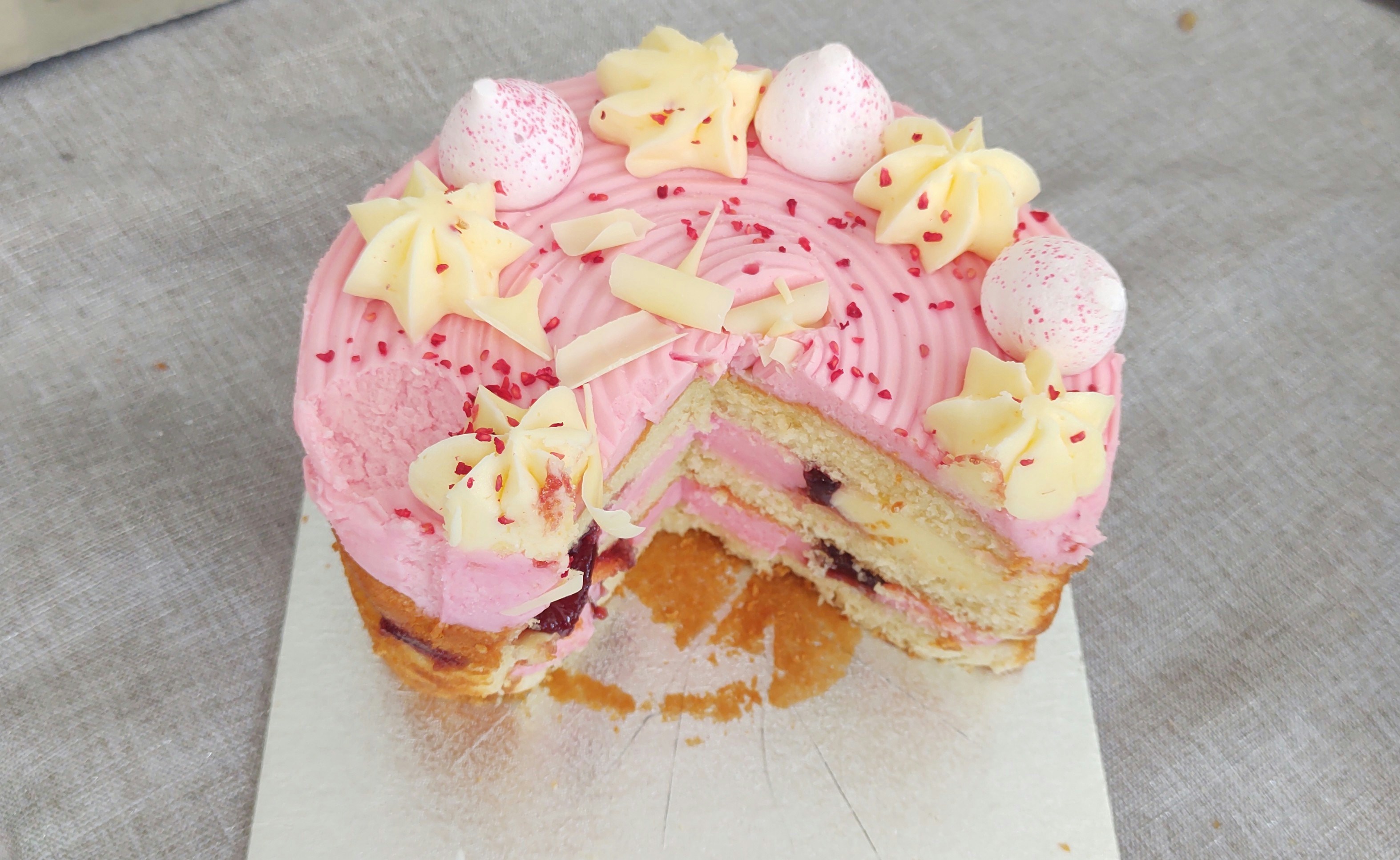 Pastel pink layered cake with decorative white chocolate petals and dollops on a white base, a single bite taken revealing creamy interior. Cake is a dessert centerpiece.