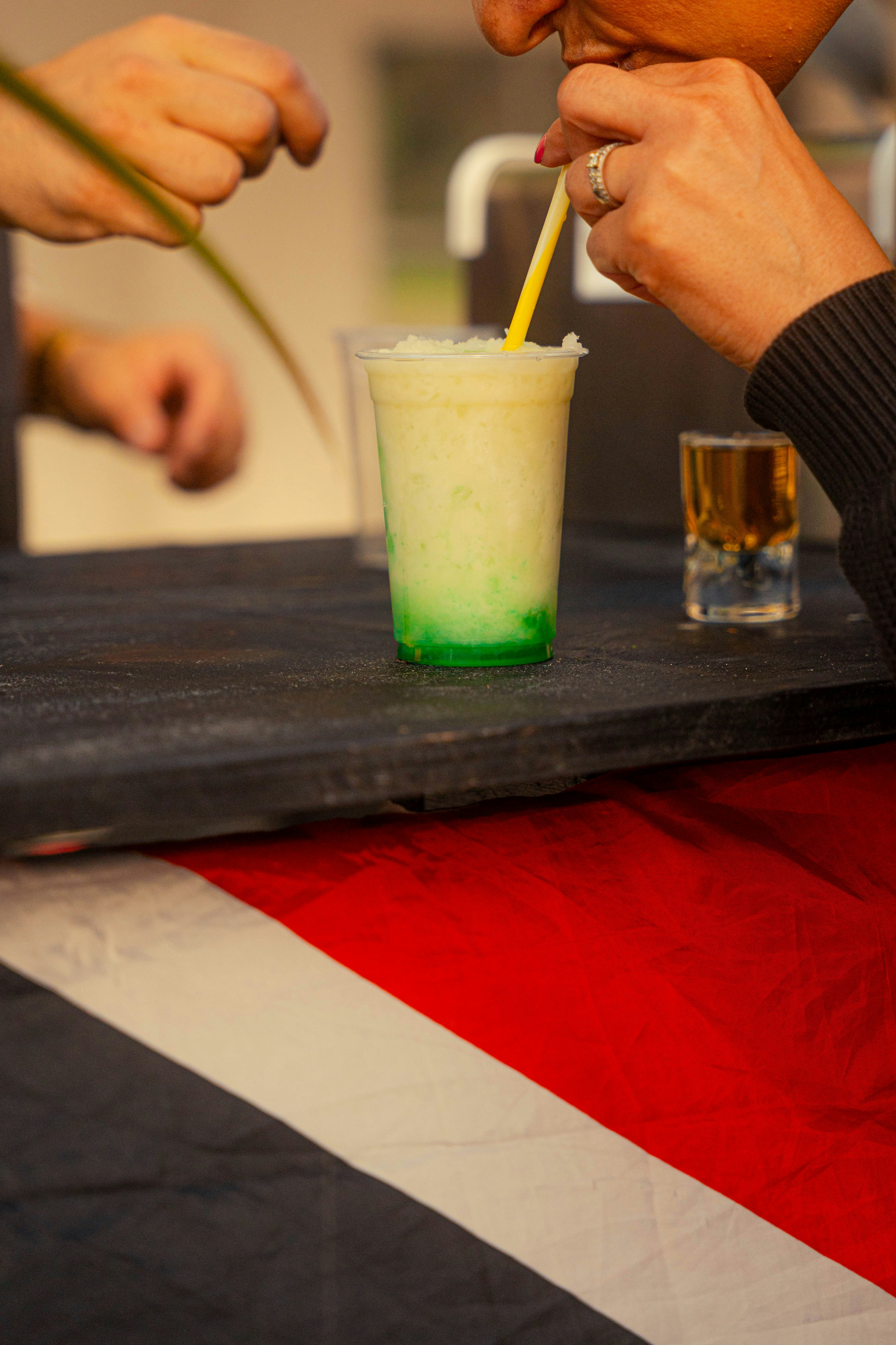 Neon-green Layered Mocktail Under Warm Bar Light, Framed by Nearby Hands