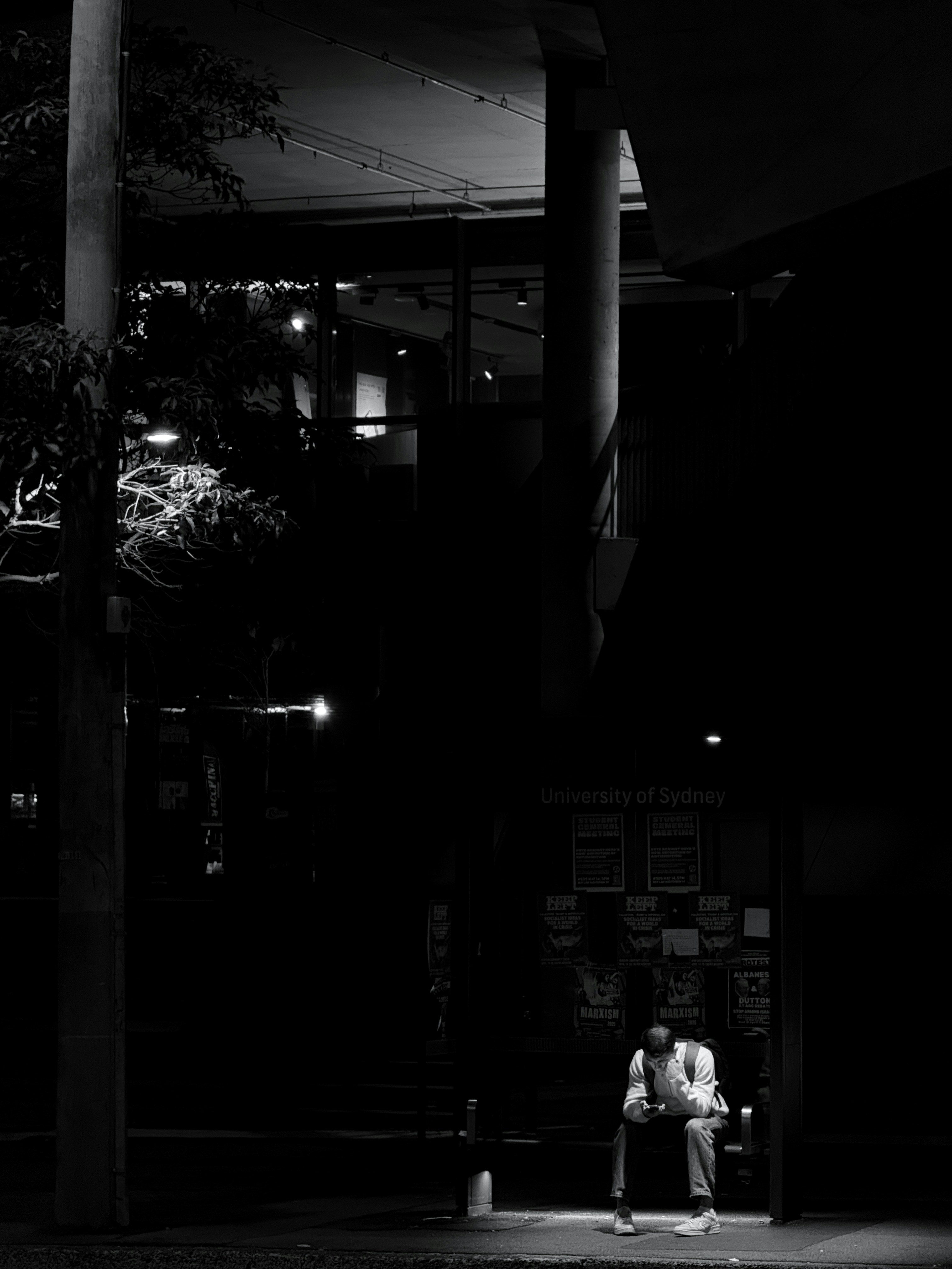 Night Watcher: A lone figure rests in a dim university plaza