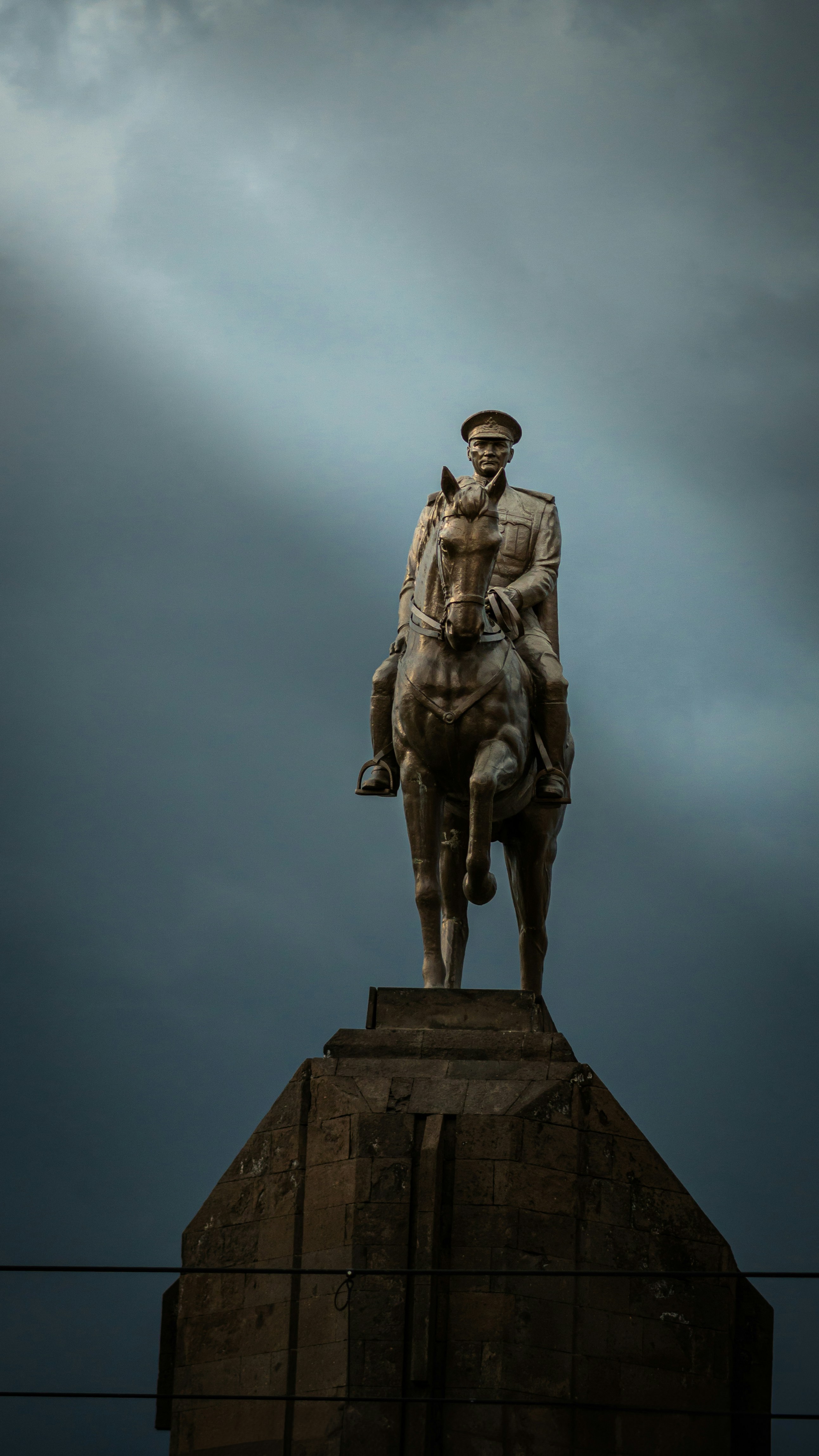 A statue of a mounted figure stands resolutely atop a stone pedestal, framed by dramatic clouds. The piece embodies strength and historical significance.
