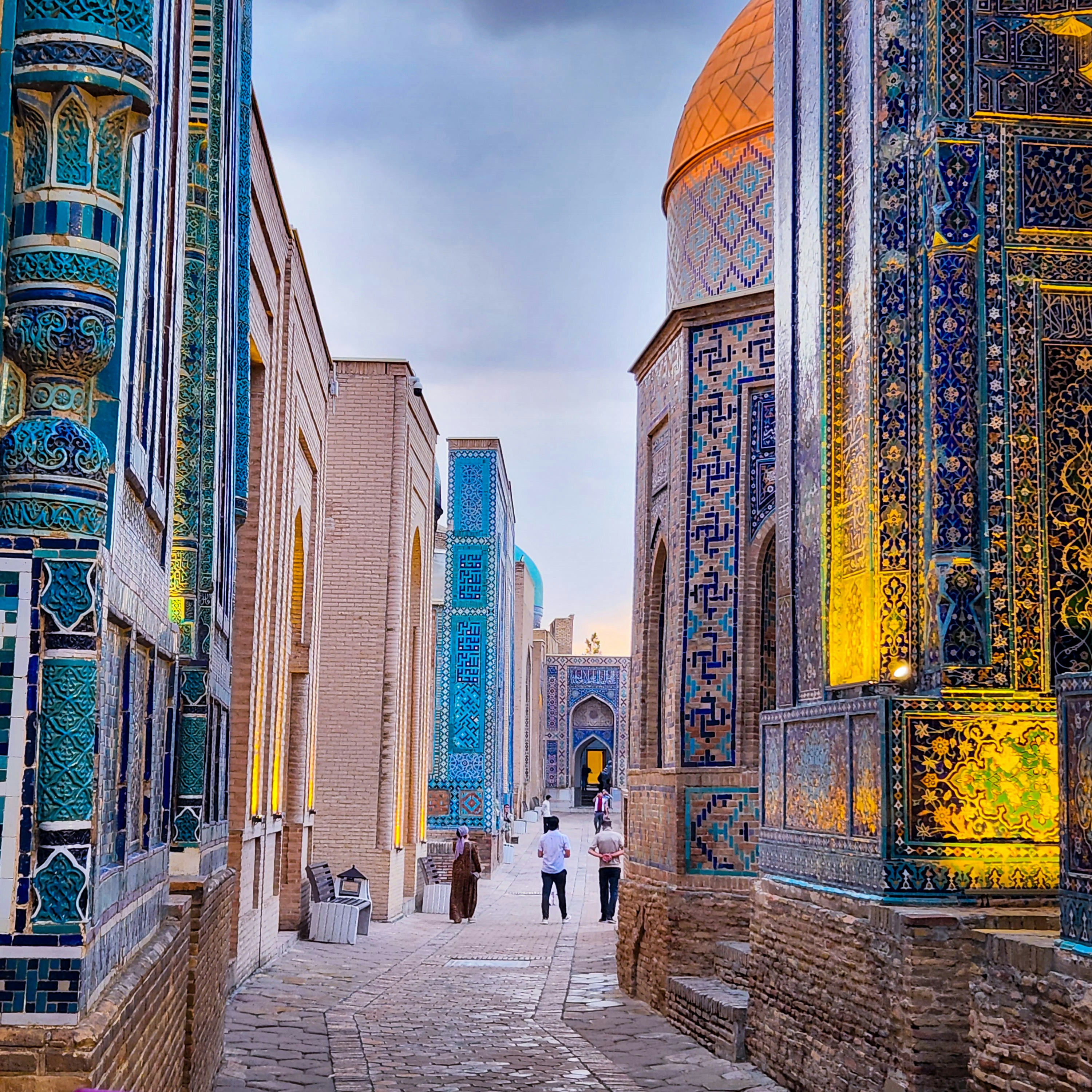 Intricate blue-tiled facades line a historic alleyway, leading to a grand entrance adorned with golden domes. Warm lights illuminate the ornate details.