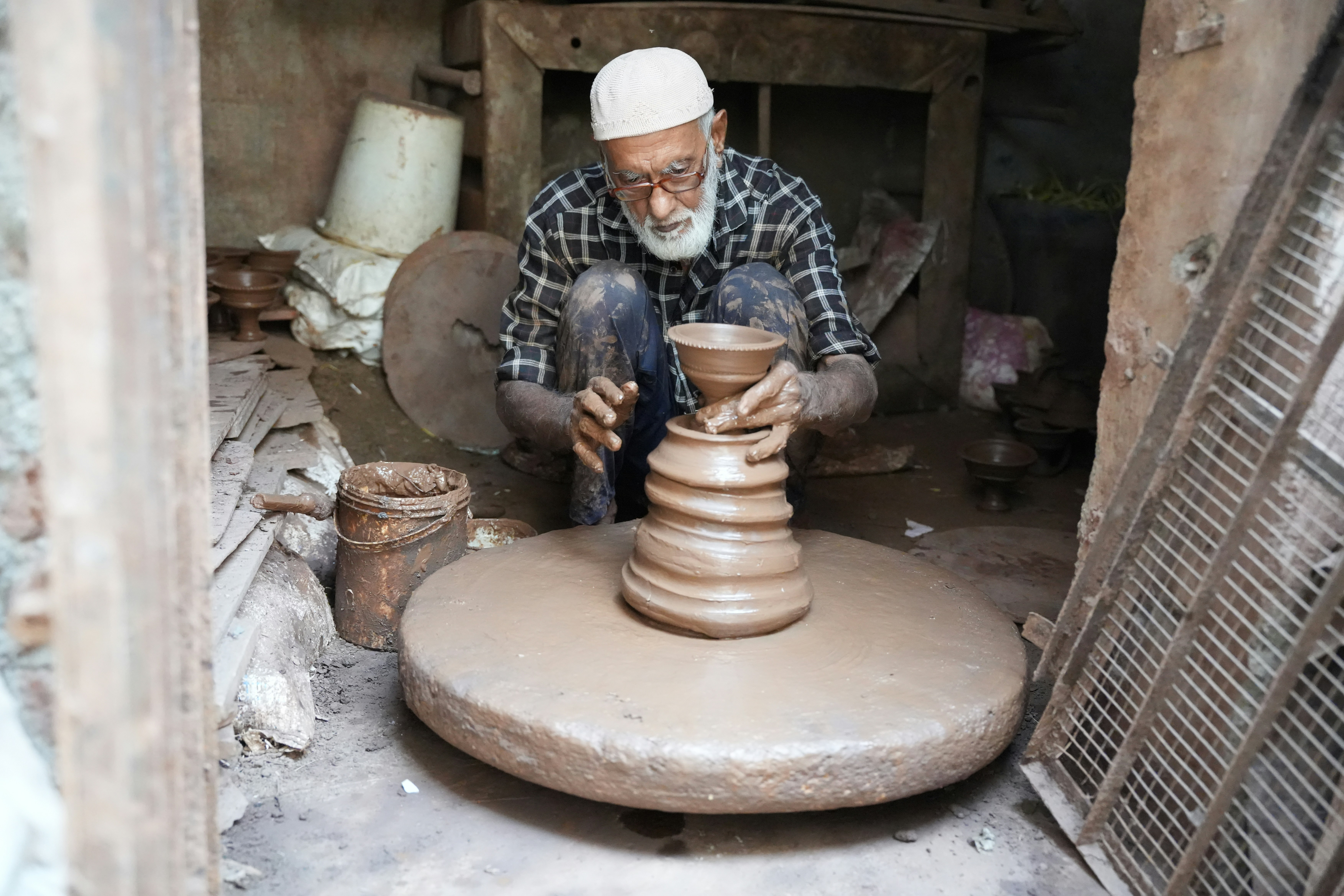 An elderly potter skillfully shaping clay on a spinning wheel in a rustic workshop, surrounded by tools and unfinished pieces.