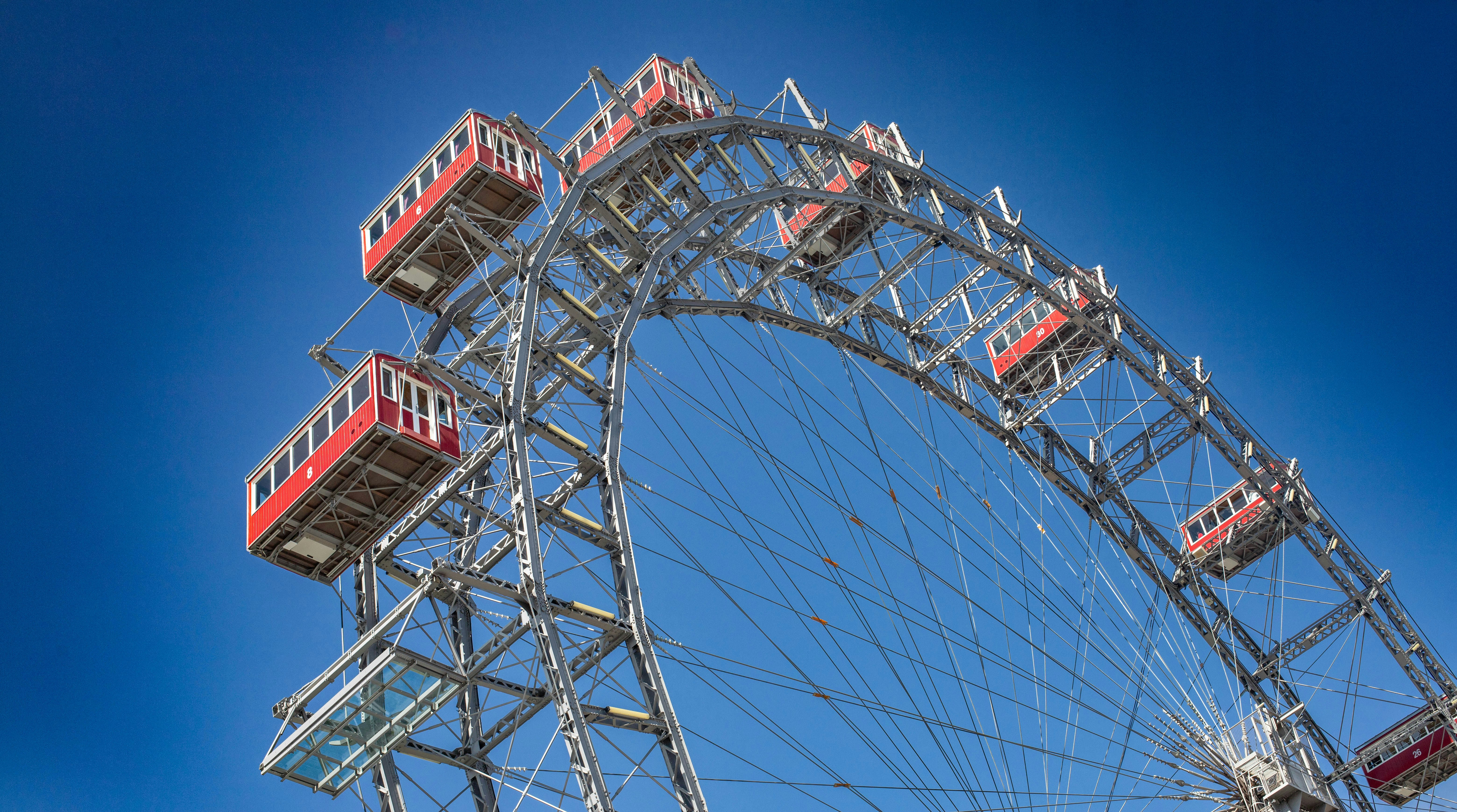 A large Ferris wheel with red rectangular cabins attached to a steel frame structure. The design includes numerous metal spokes connecting the cabins to the central axis. The wheel is photographed from different angles and lighting conditions, showing its architectural details and vertical orientation. The structure is part of a well-known amusement park attraction. | A ferris wheel against a bright blue sky.