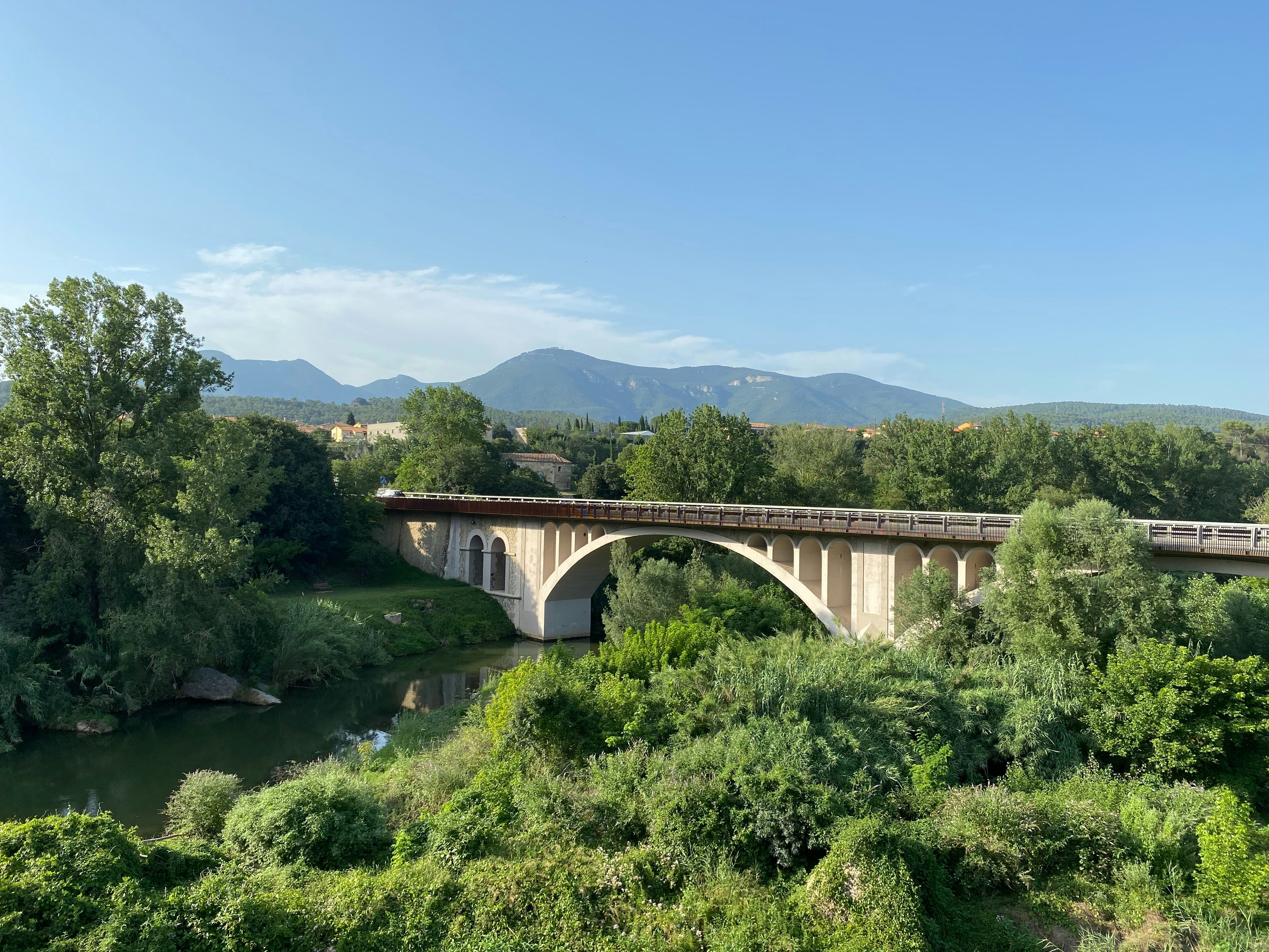 A picturesque bridge gracefully arches over a tranquil river, framed by lush greenery and distant mountains. The scene captures the harmony of nature and infrastructure.