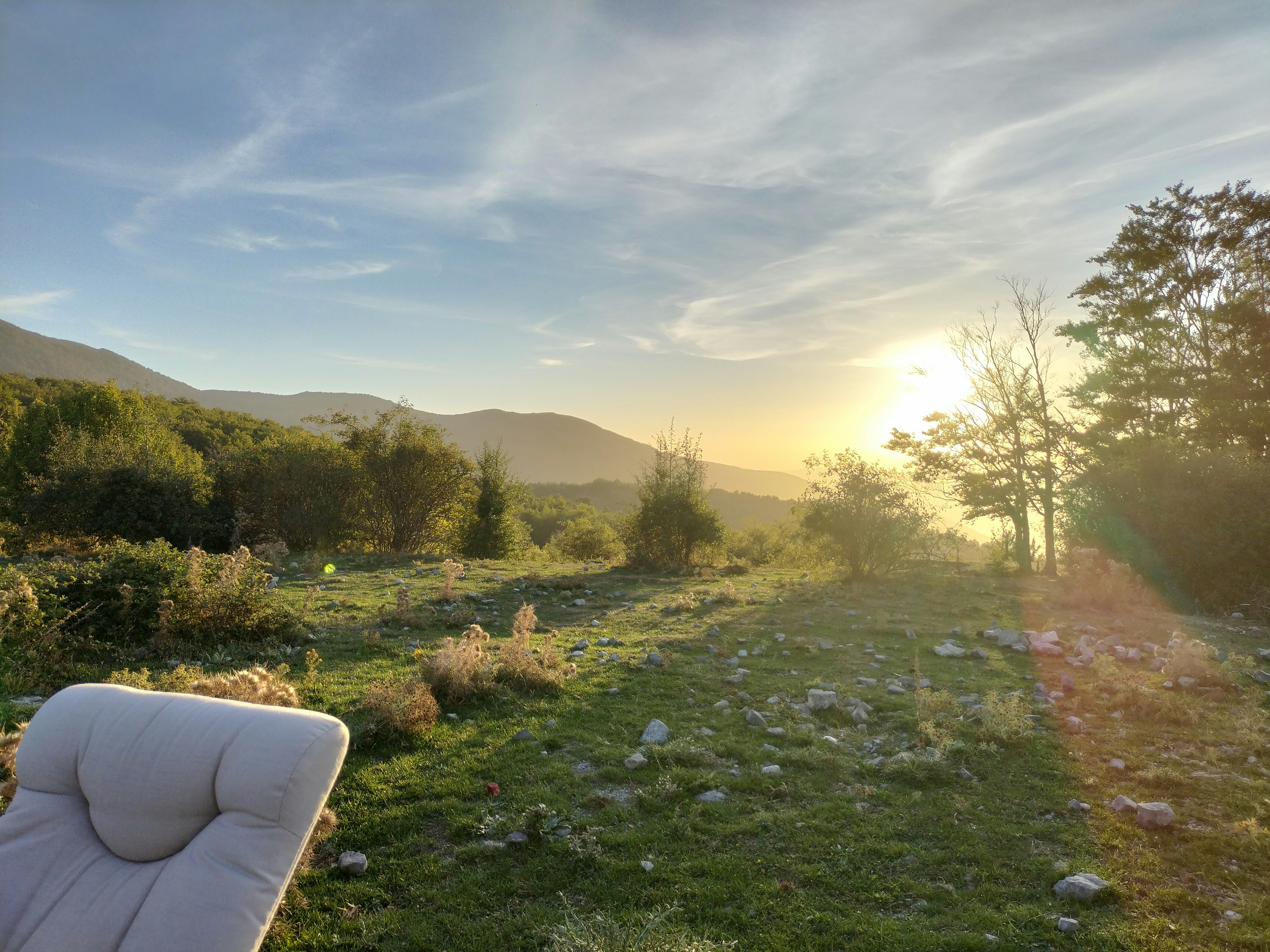 A photograph captures a sunlit meadow with a lone cushioned chair in the foreground as the sun dips behind distant hills.