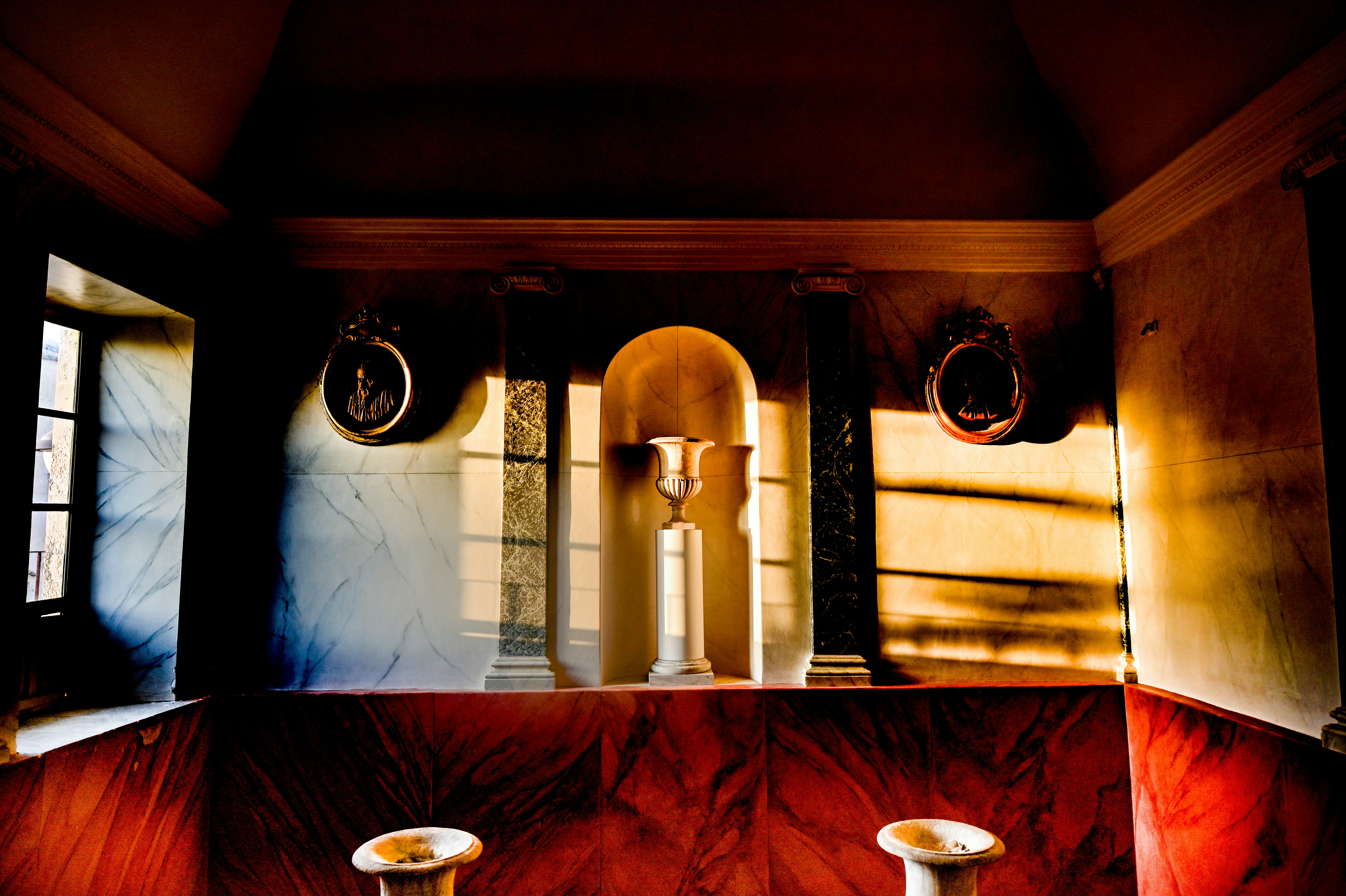 Sunlight casts intricate shadows across a marble chamber, highlighting a central pedestal and ornate decorations on the walls.