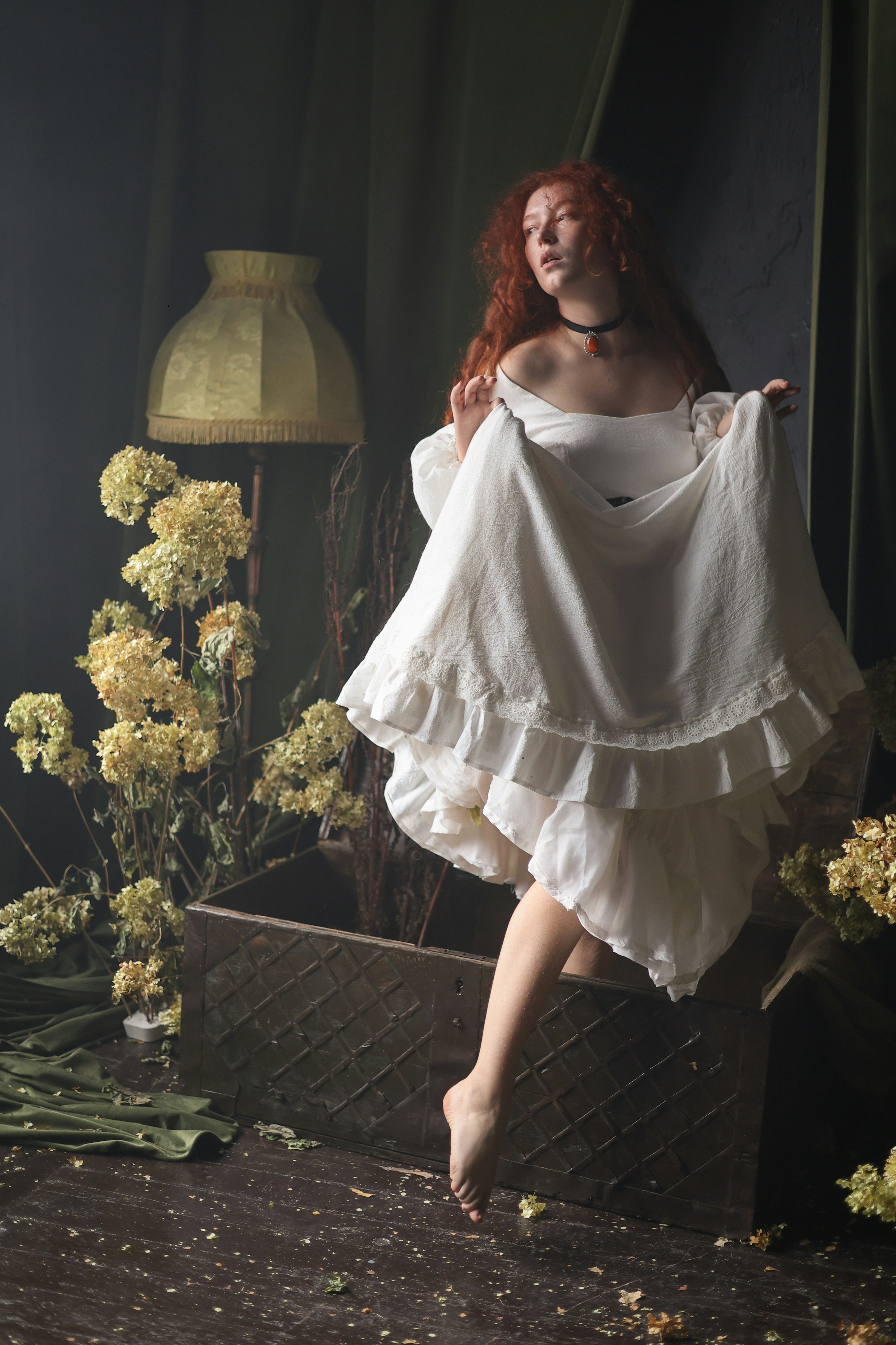 A woman in a flowing white dress gracefully poses amidst dried flowers and soft fabrics, evoking a sense of nostalgia and tranquility.