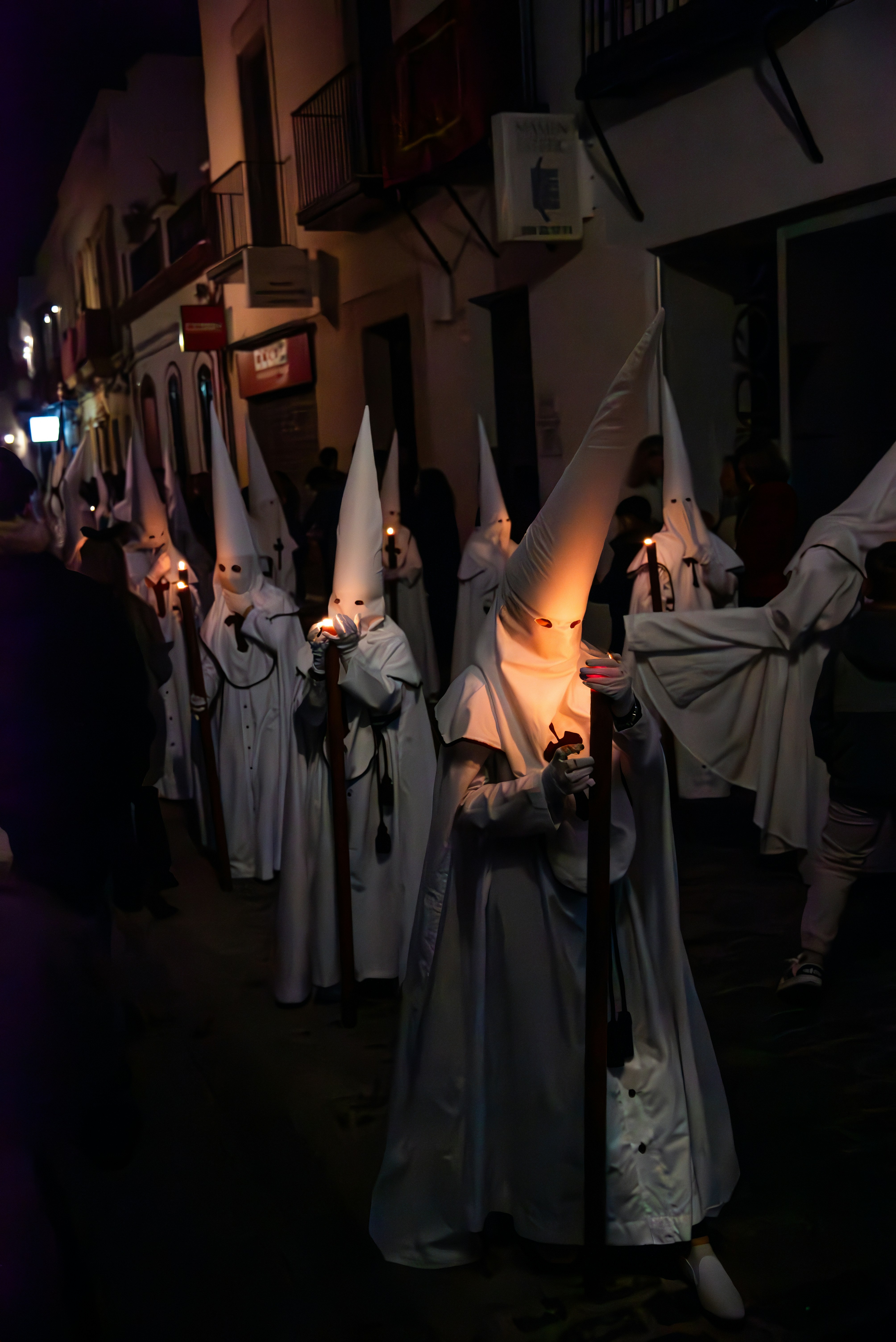 A procession of hooded figures carrying candles through a narrow street, cloaked in white robes and pointed hats, creating an atmosphere of mystery and reverence.