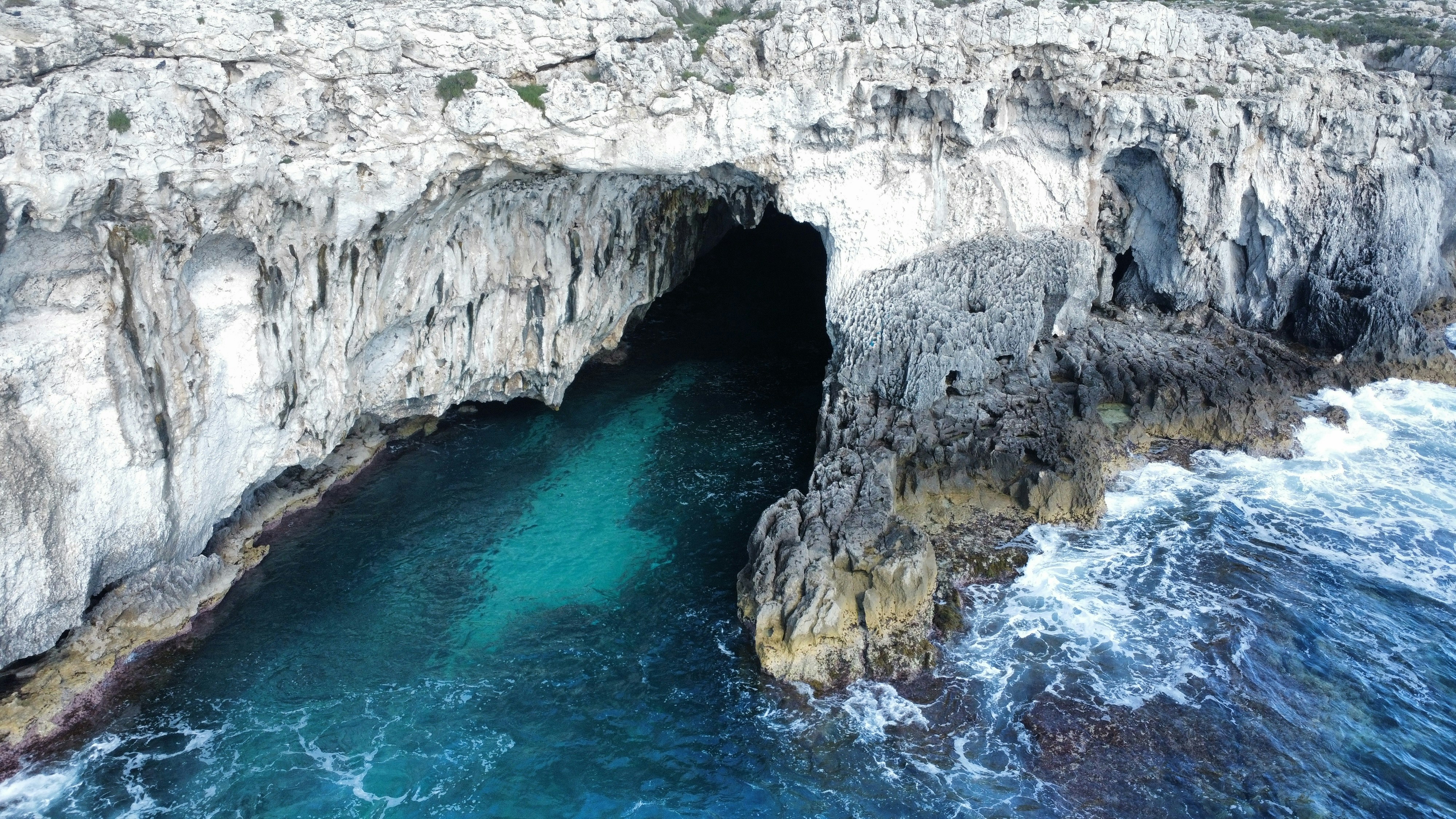 A dramatic coastal cave entrance reveals turquoise waters contrasting with rugged cliffs. The scene captures the dynamic interplay between land and sea.