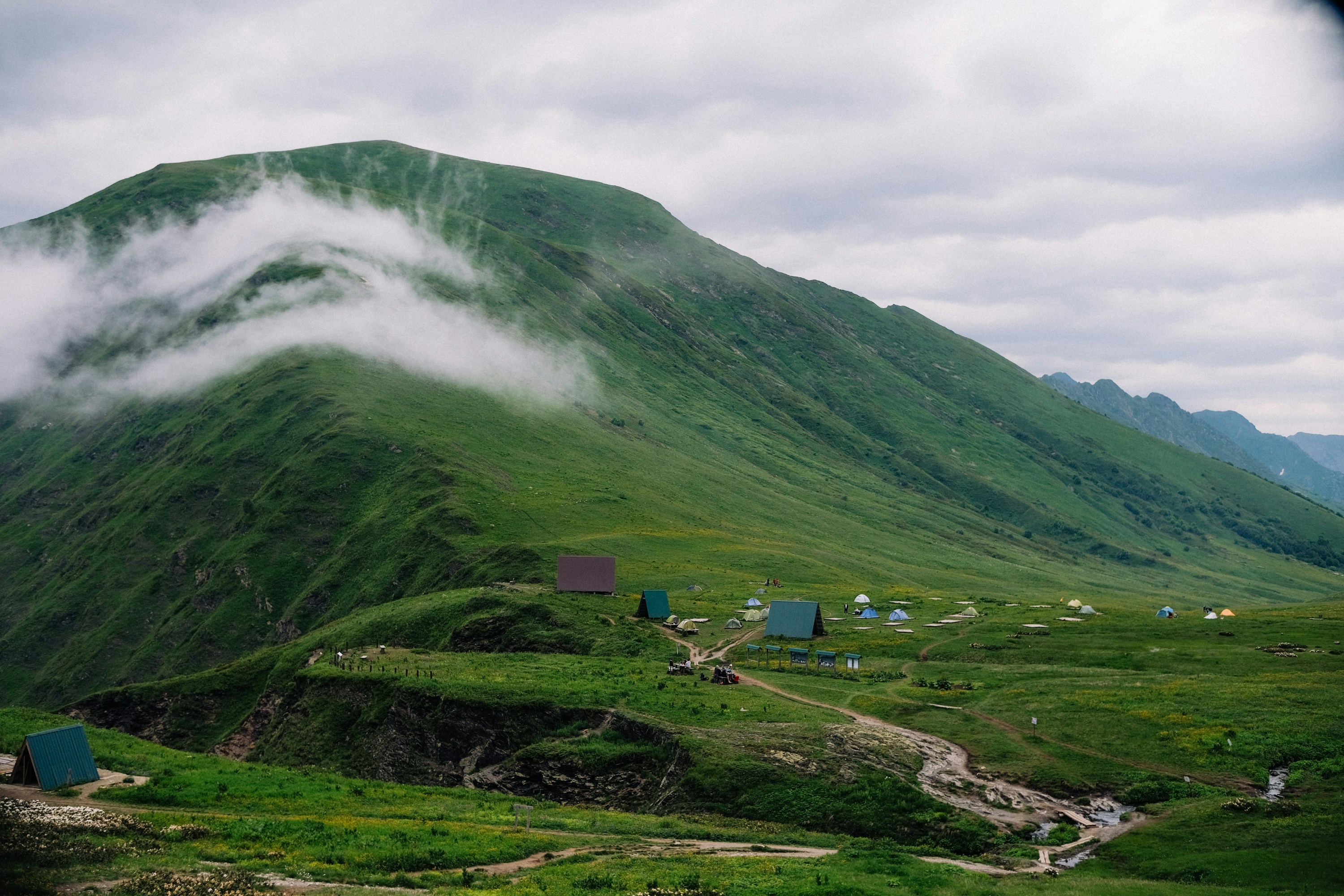 Lush green hills envelop quaint camping structures under a cloudy sky, showcasing the harmony of nature and human presence.