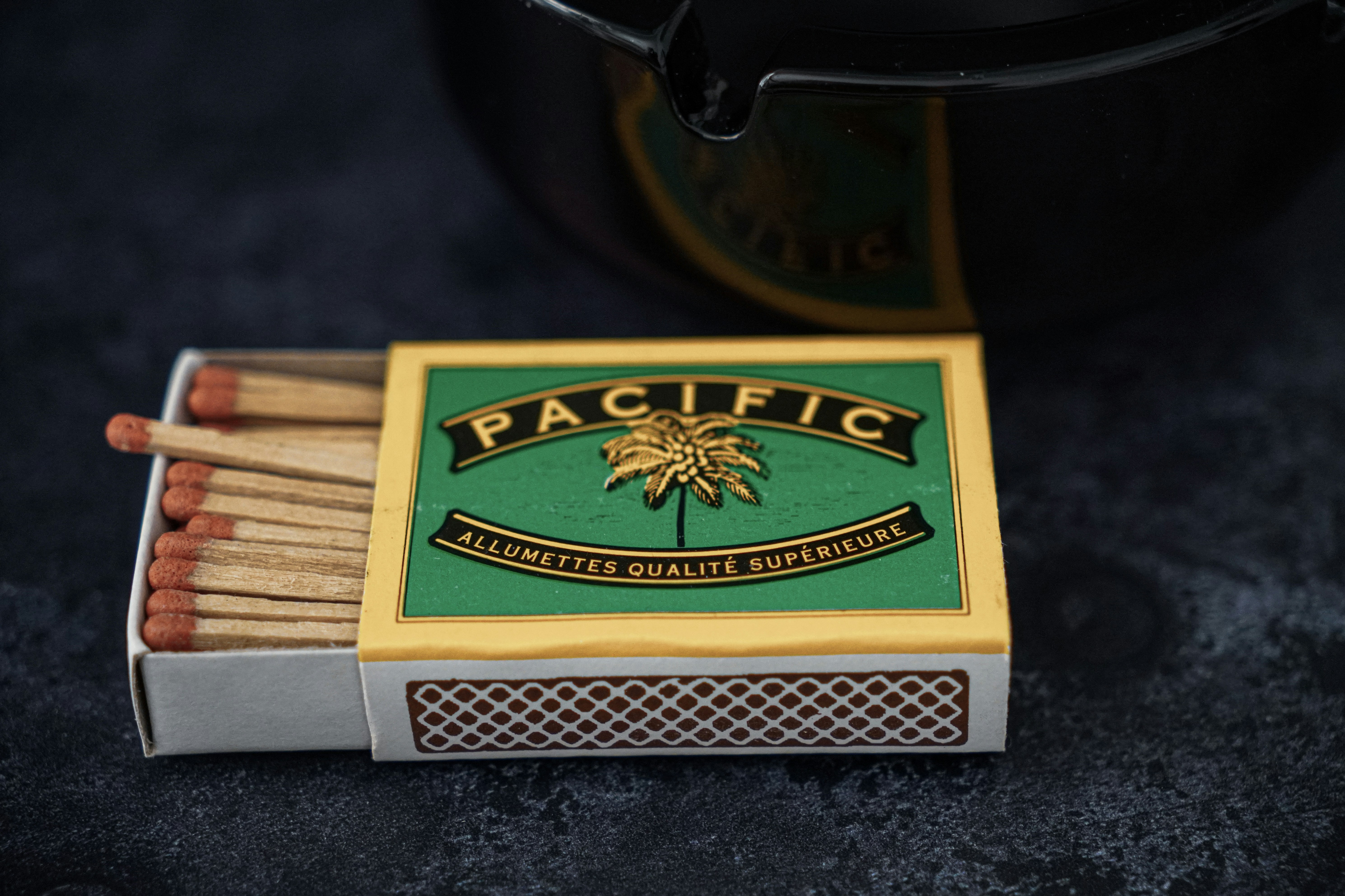 Close-up of a Pacific matchbox logo on a box of matches resting on a dark surface. A few wooden matches protrude from the side, forming a quiet still life.