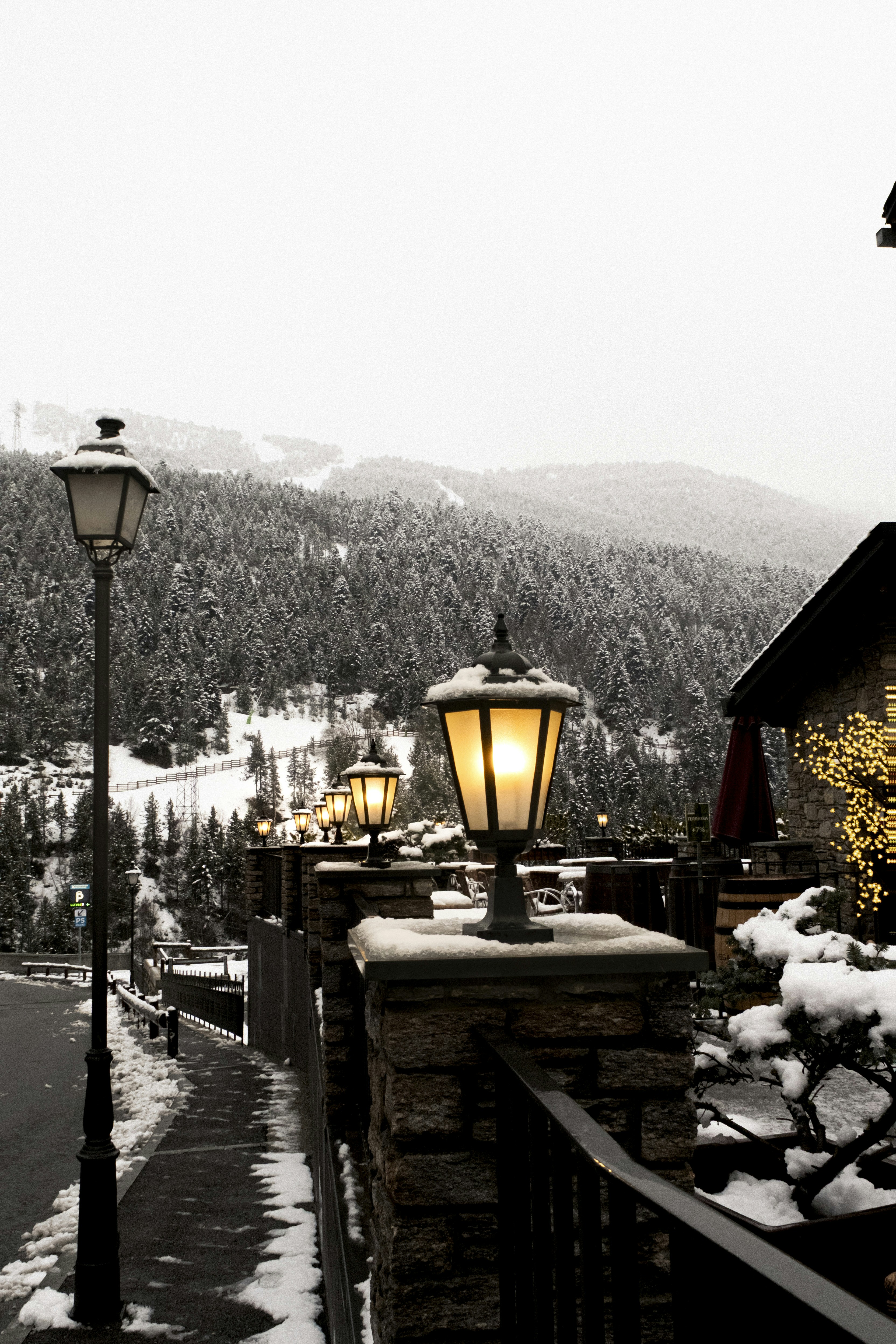 Snow-covered street adorned with vintage lanterns illuminating a serene winter landscape, framed by evergreen trees and a distant mountain range.