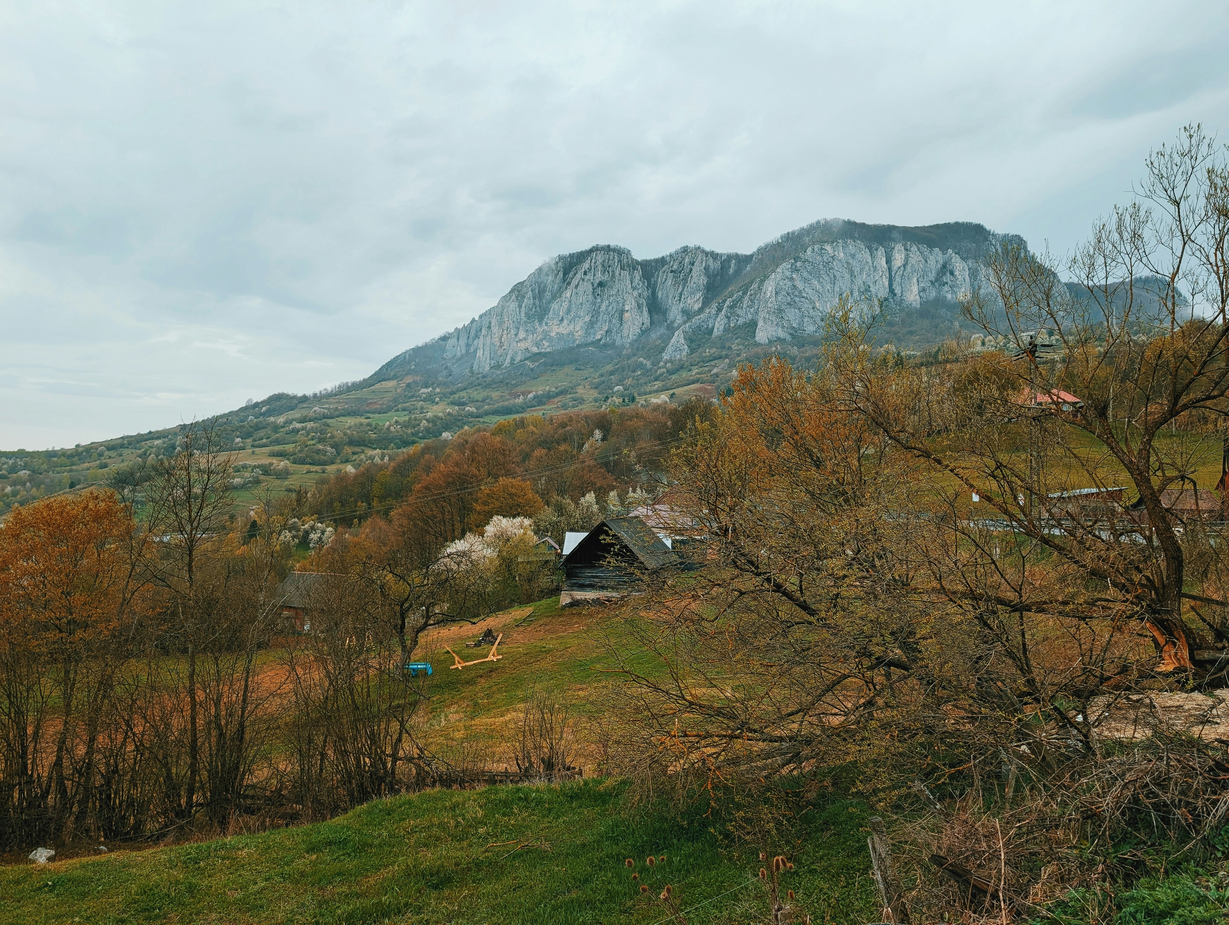 Autumn landscape featuring rolling hills, a rustic cabin, and towering mountains under a cloudy sky.