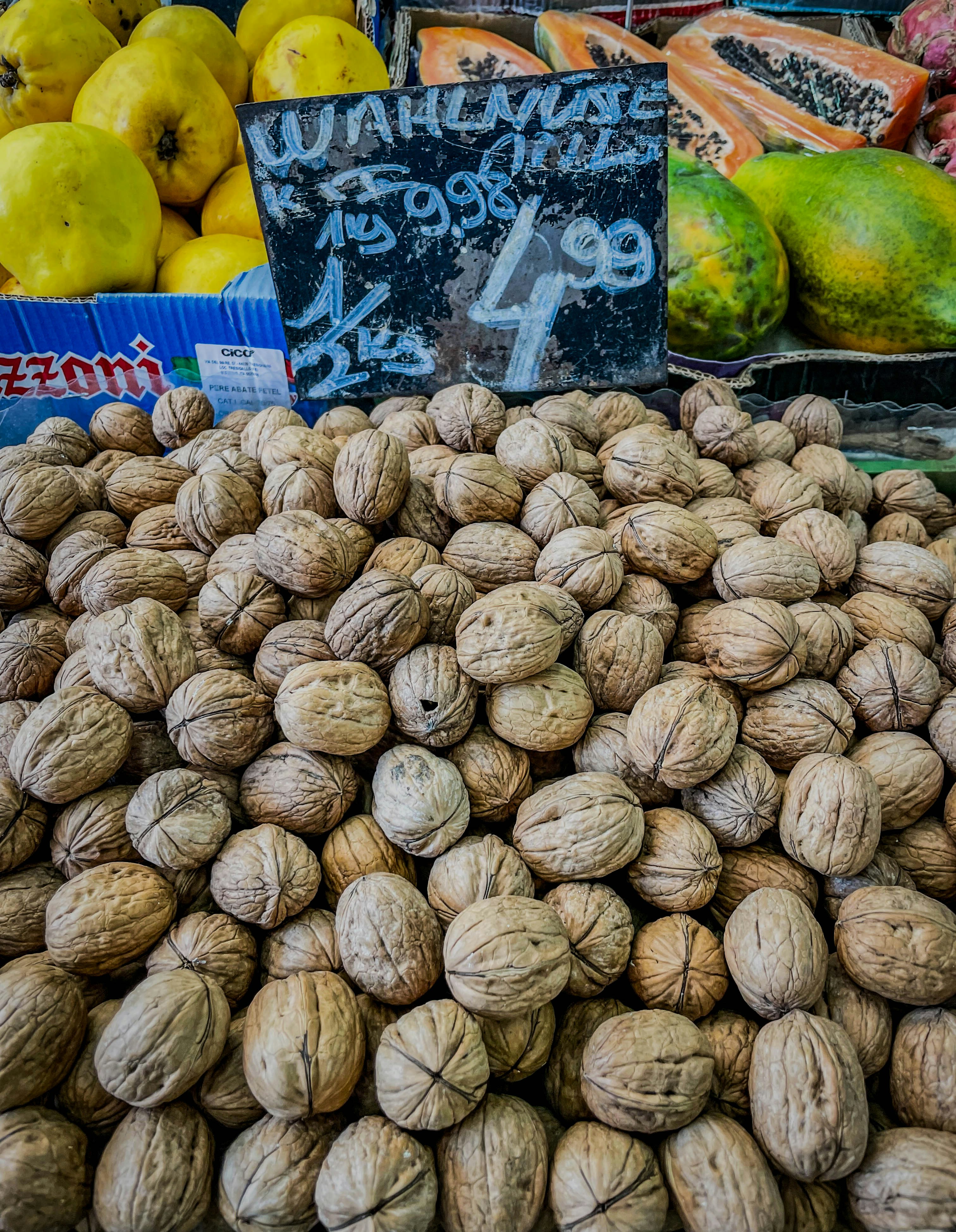A market stall displays a large pile of unshelled walnuts in the foreground, with various tropical fruits such as papayas, quinces, and dragon fruit arranged in the background. A handwritten chalkboard sign indicates pricing information.