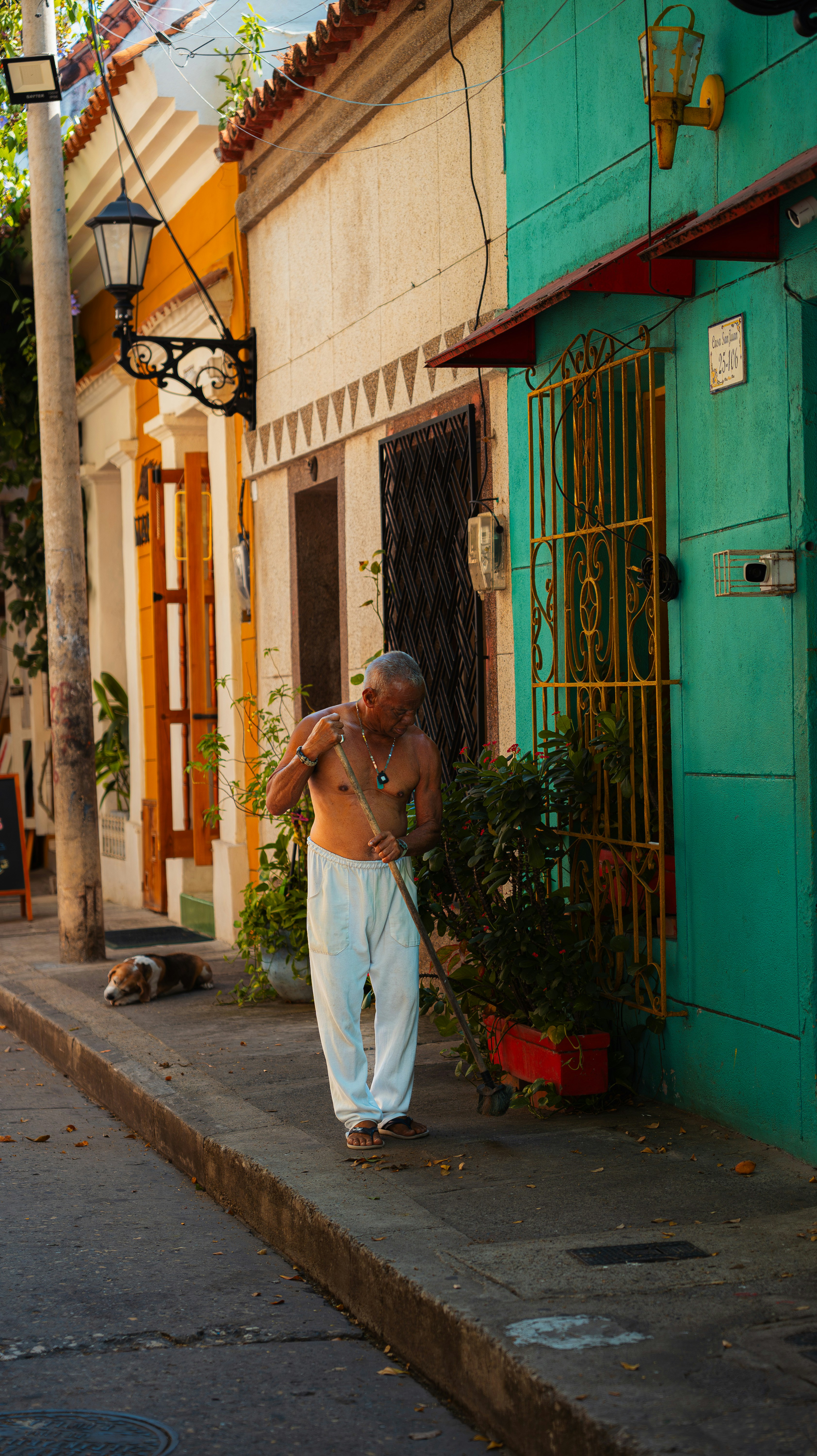 Elderly man sweeping the sidewalk in a vibrant neighborhood, with a relaxed dog resting nearby. The scene captures daily life in a charming locale.