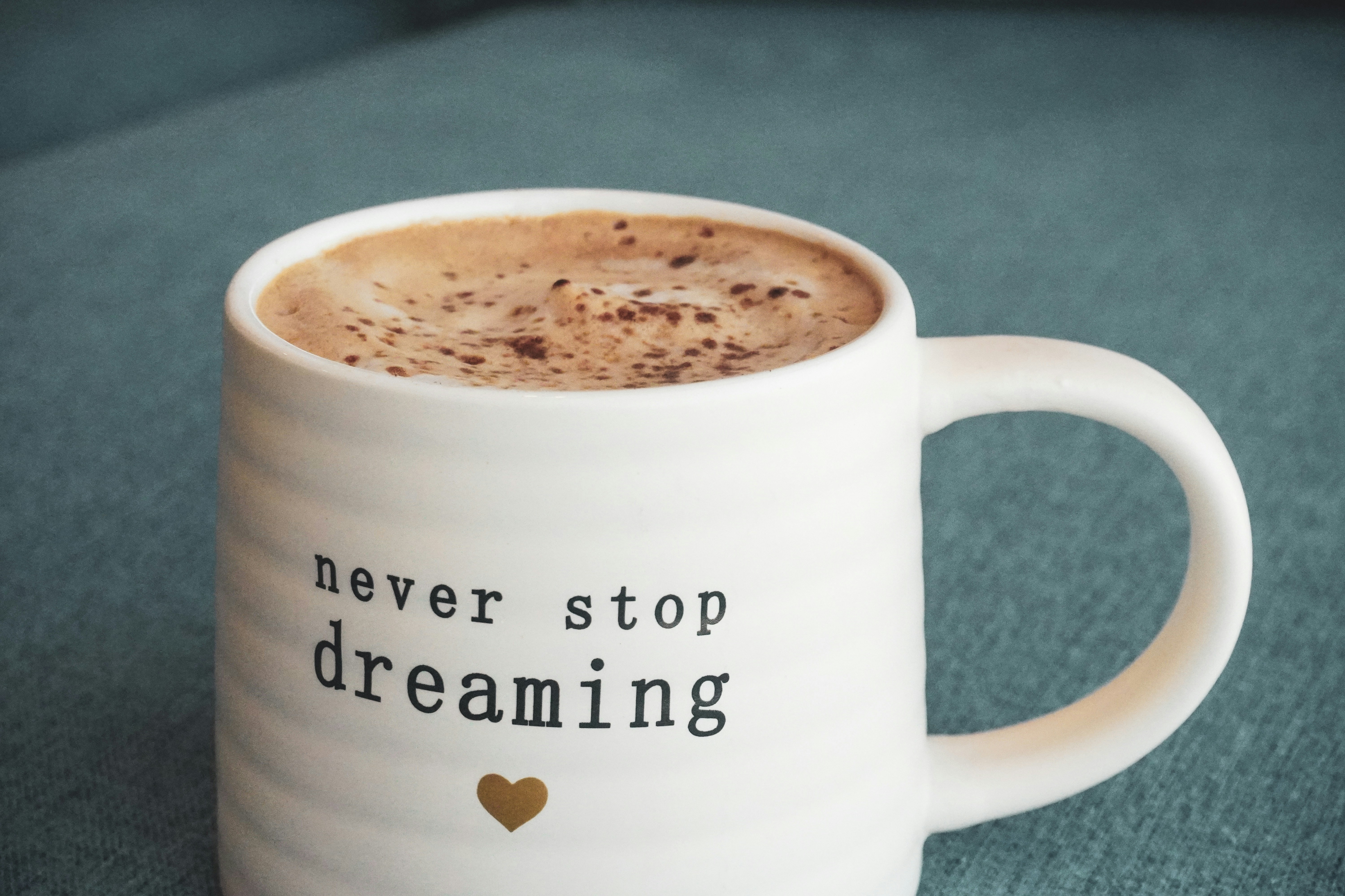 Ceramic mug with the phrase 'never stop dreaming' and a heart, filled with frothy coffee. The mug rests on a soft, textured surface.