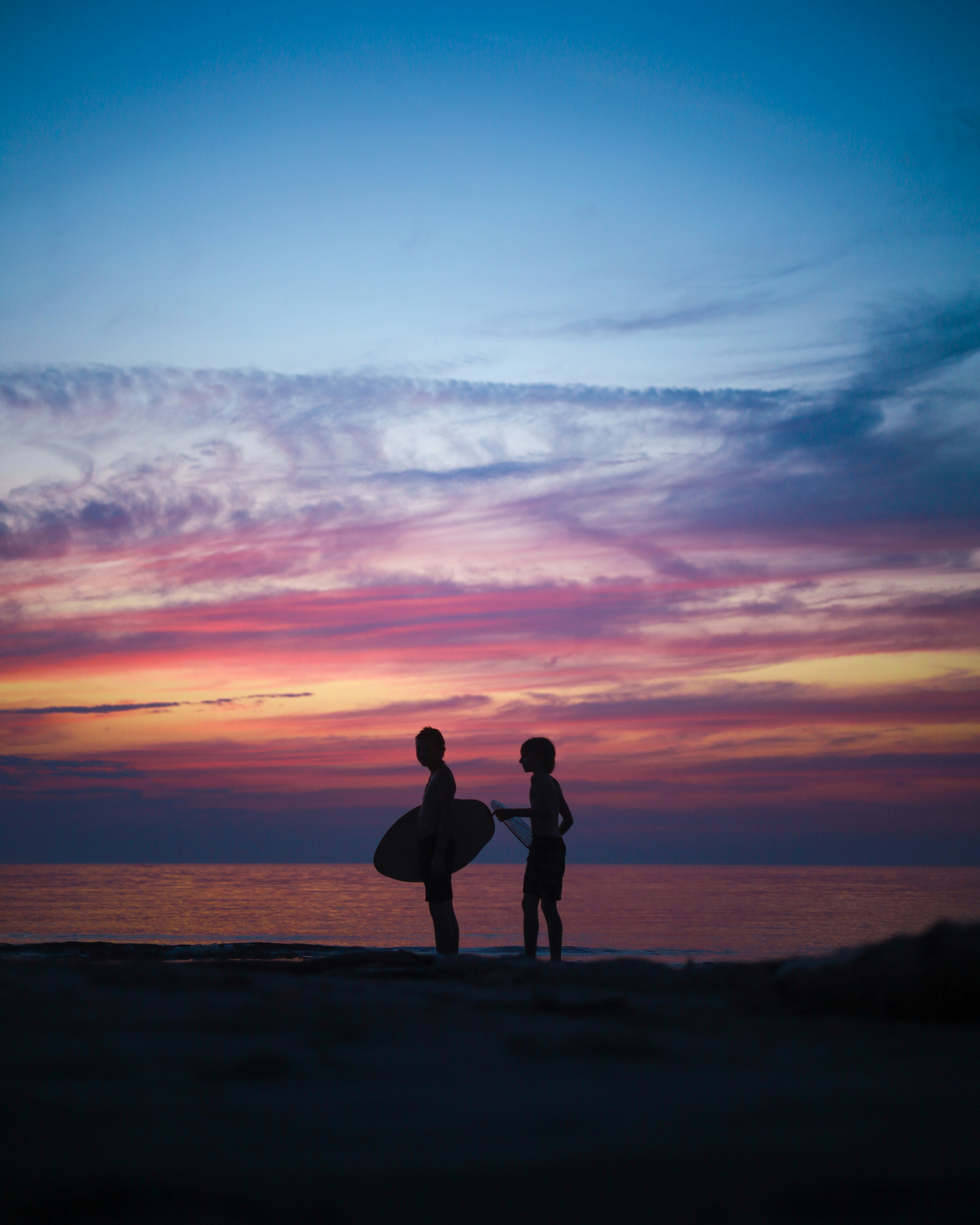 Two silhouetted children converse by the ocean at sunset, one holding a surfboard. The vibrant sky transitions from blue to warm hues of orange and pink.