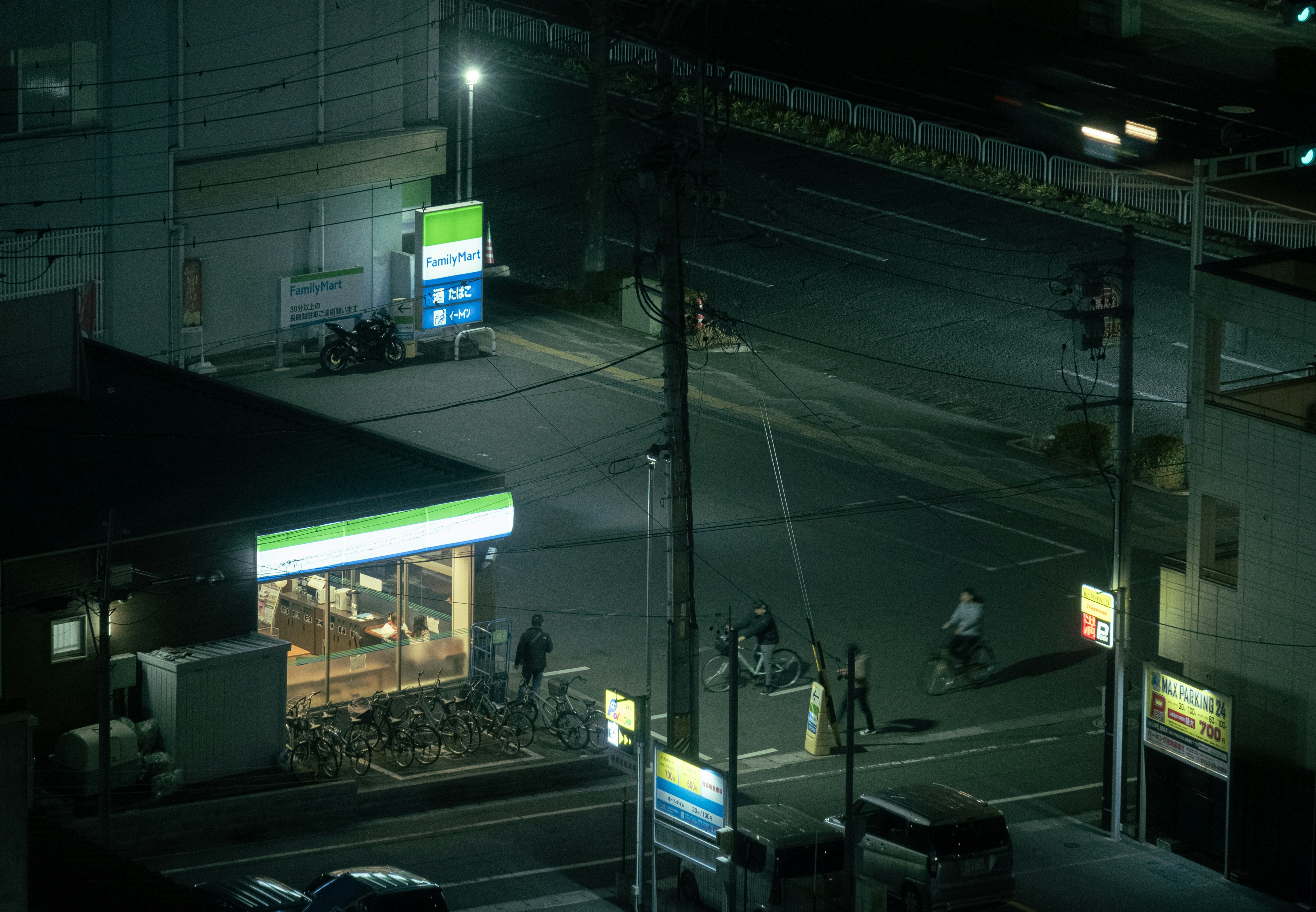 A quiet urban scene at night featuring a convenience store illuminated by soft light, with cyclists navigating the street. The atmosphere captures the essence of city life after dark.