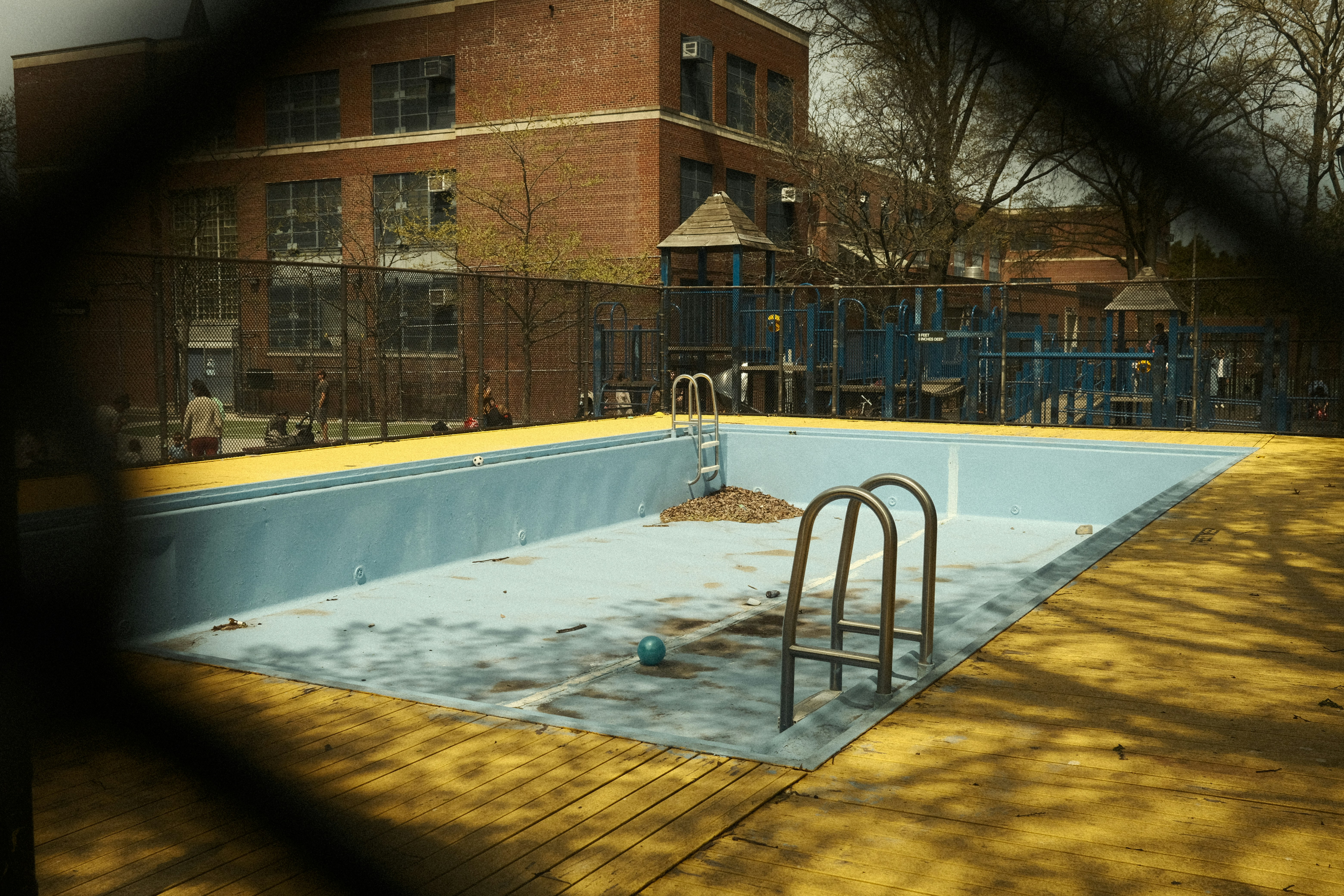 Empty swimming pool surrounded by a chain-link fence, with playground equipment in the background. The scene evokes a sense of nostalgia and abandonment.