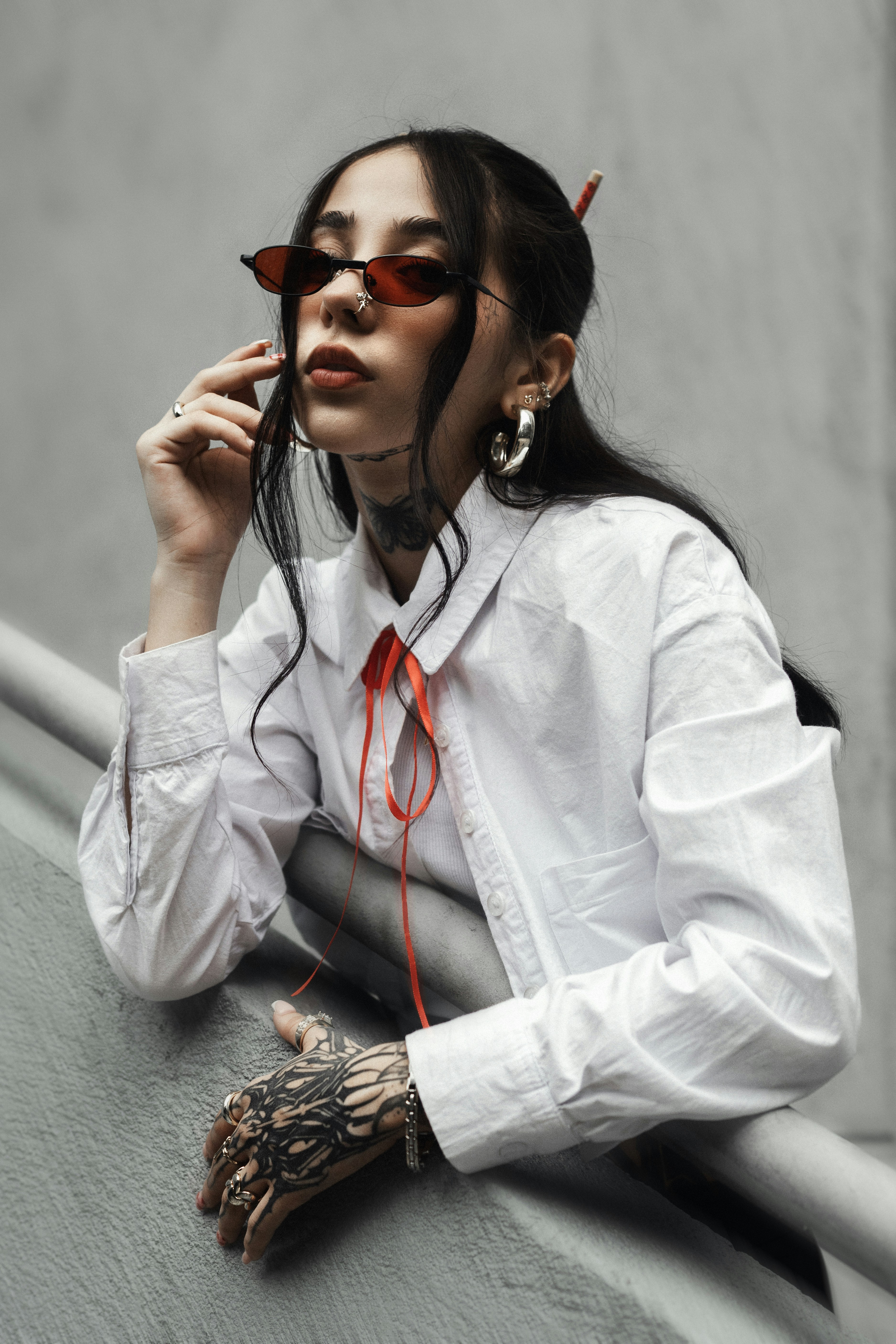 Photographic portrait of a woman in a white shirt with red accents, leaning on a concrete railing against a gray urban backdrop.