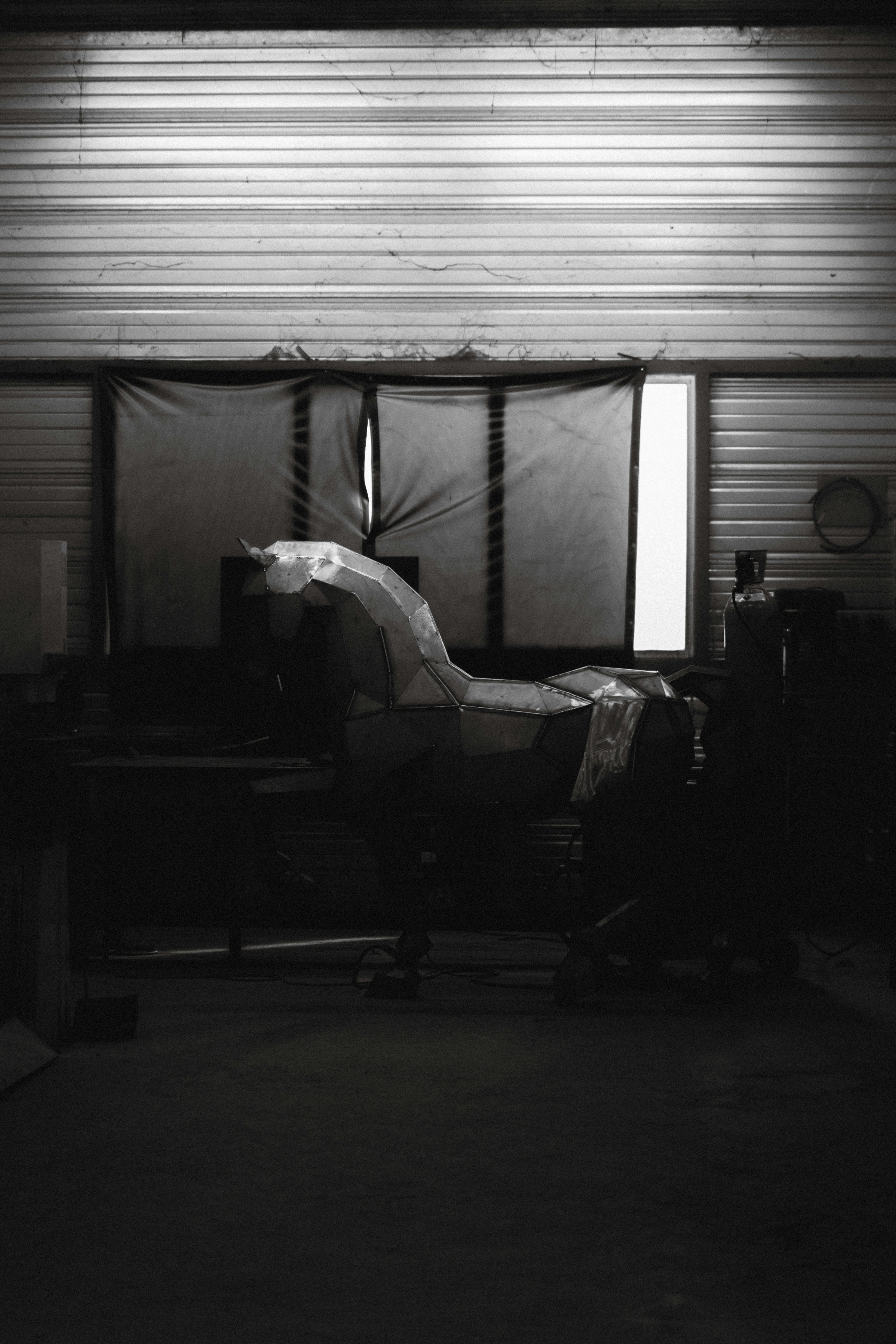 Geometric horse sculpture stands in dimly lit workshop, surrounded by shadows and industrial elements.