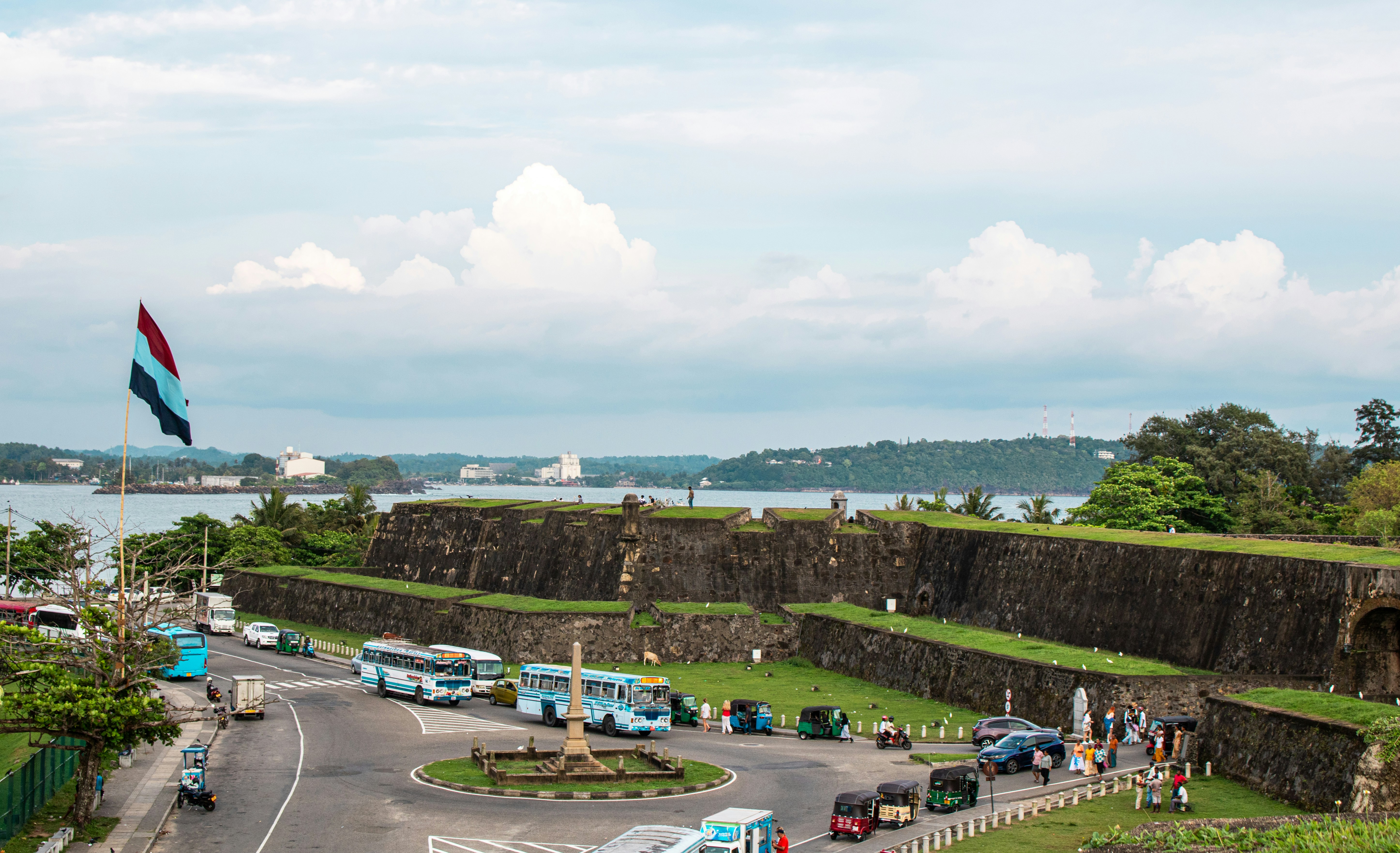 A busy traffic circle operates beside the historic stone ramparts of Galle Fort, Sri Lanka. Colorful public buses (Ashok Leyland models) and ubiquitous tuk-tuks navigate the junction around a central monument.