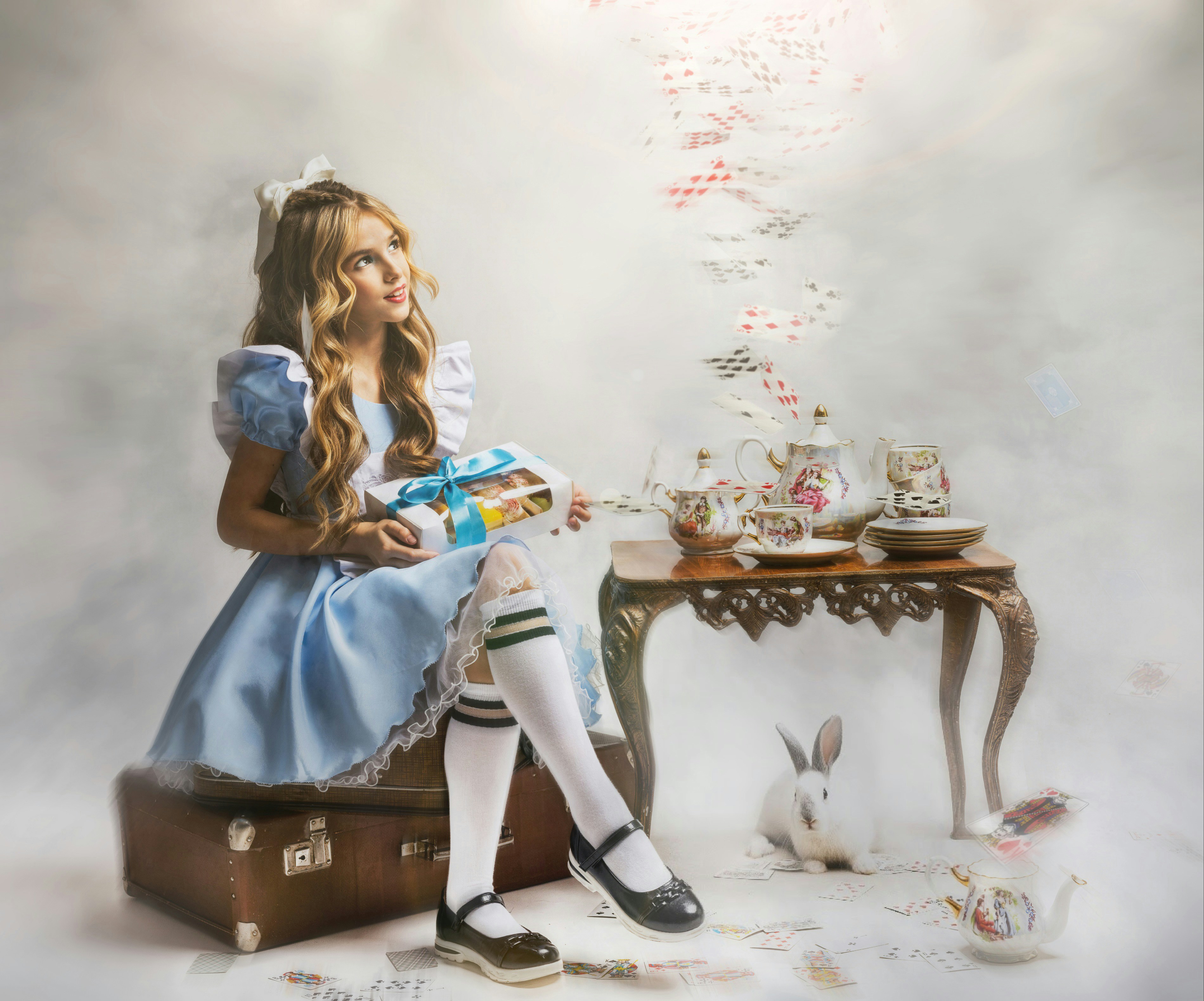 Young girl in a blue dress sits on a suitcase, holding a gift, surrounded by a whimsical tea set and a white rabbit. Playing cards float around her, evoking a sense of wonder.