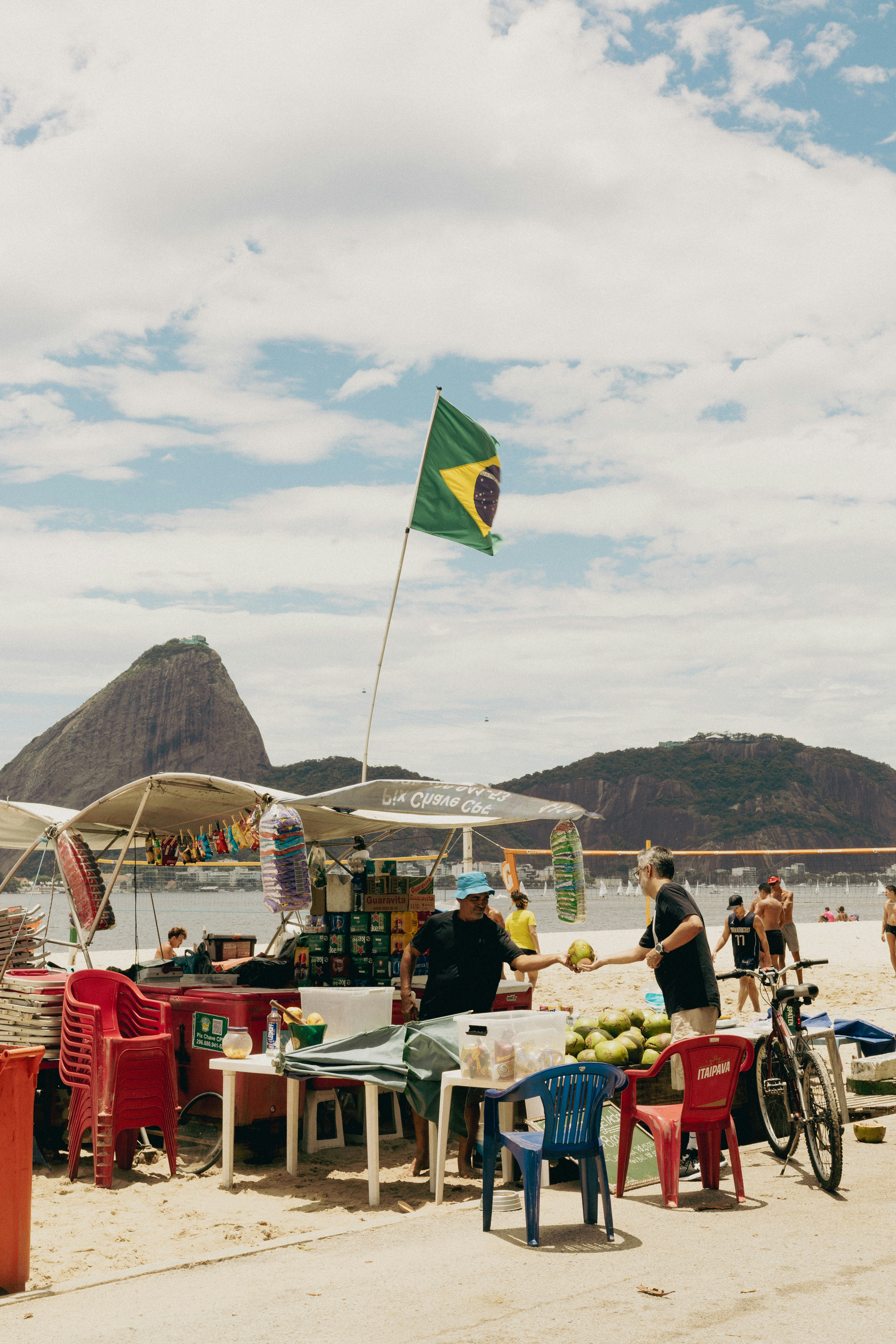 Beach market stall on a sandy shore with a tall Brazilian flag, colorful chairs, and vendors, with rocky hills in the distance.