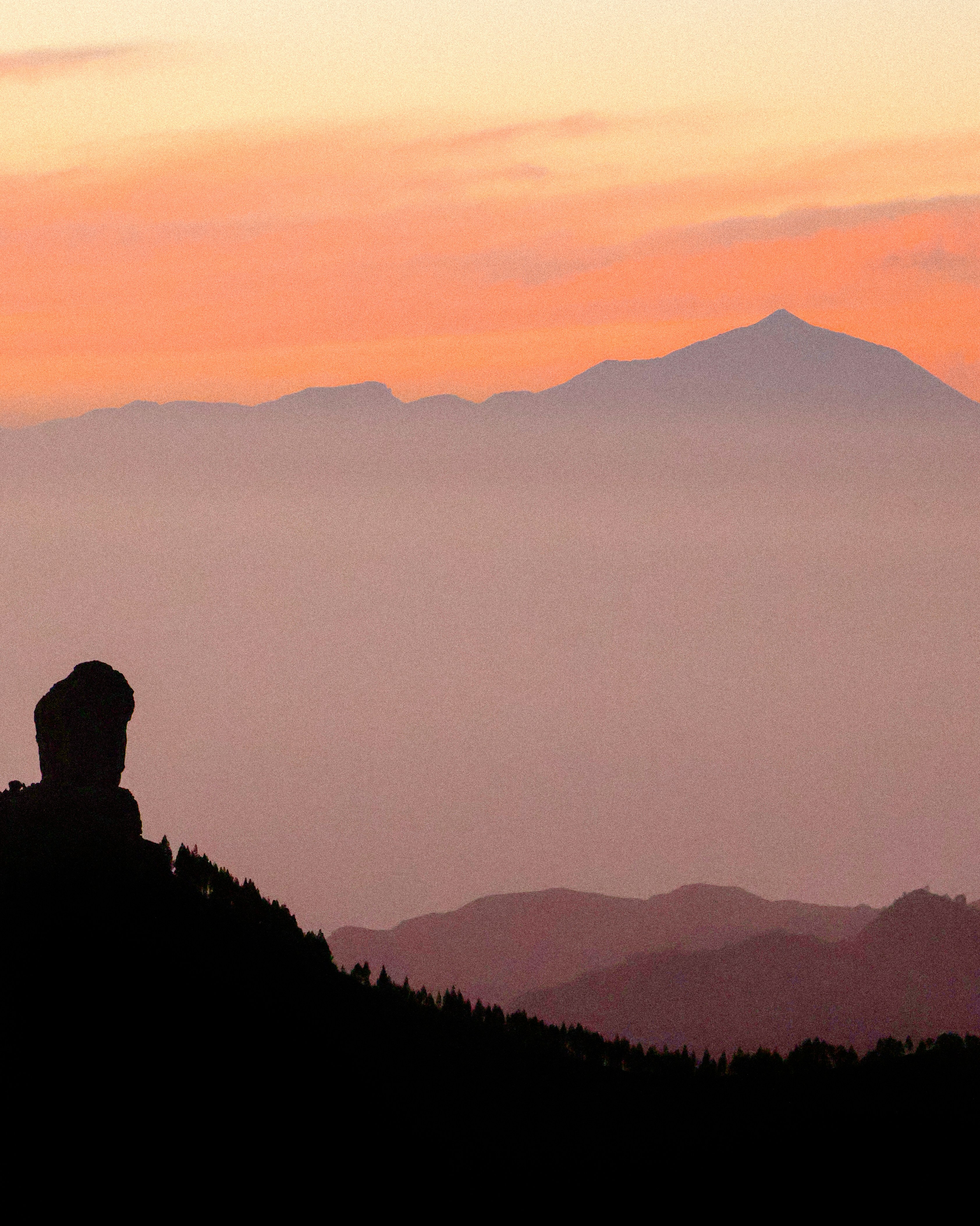 A solitary rock formation stands in silhouette against a pastel sky, with distant mountains fading into the horizon. The scene captures the serene transition of day to night.