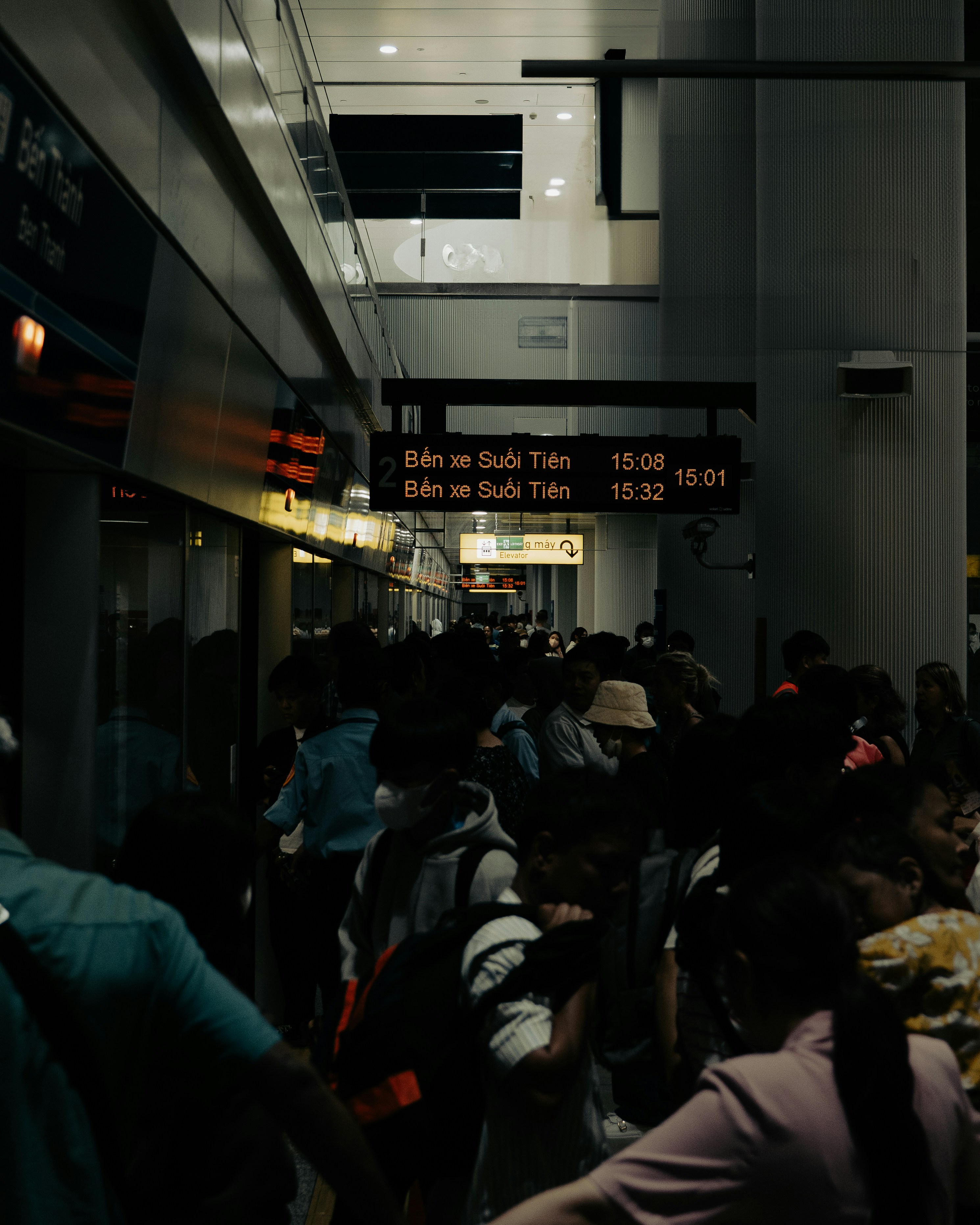 Crowd of commuters waiting at a bustling subway station with digital schedule displays overhead.